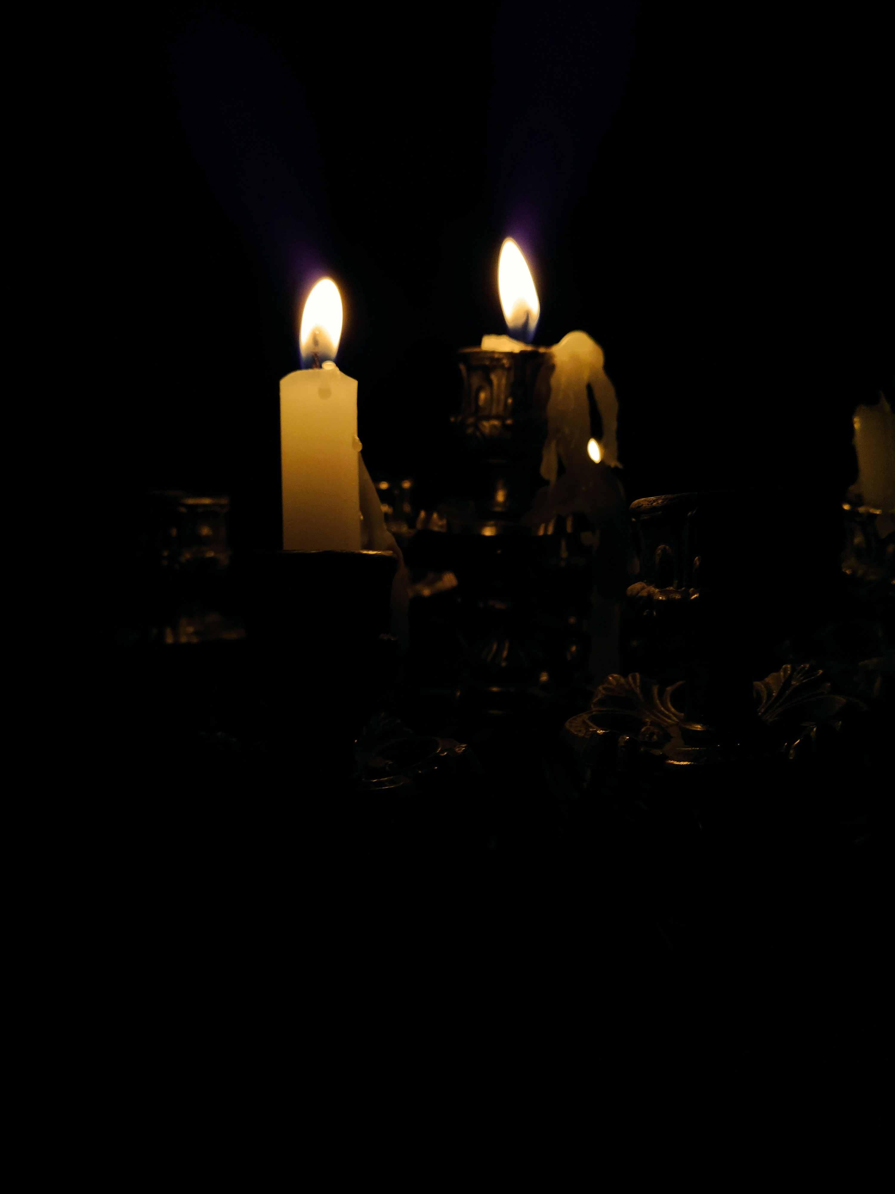 Candles flicker softly in a dimly lit setting, casting a warm glow amid shadows. The scene evokes a sense of tranquility and reflection.