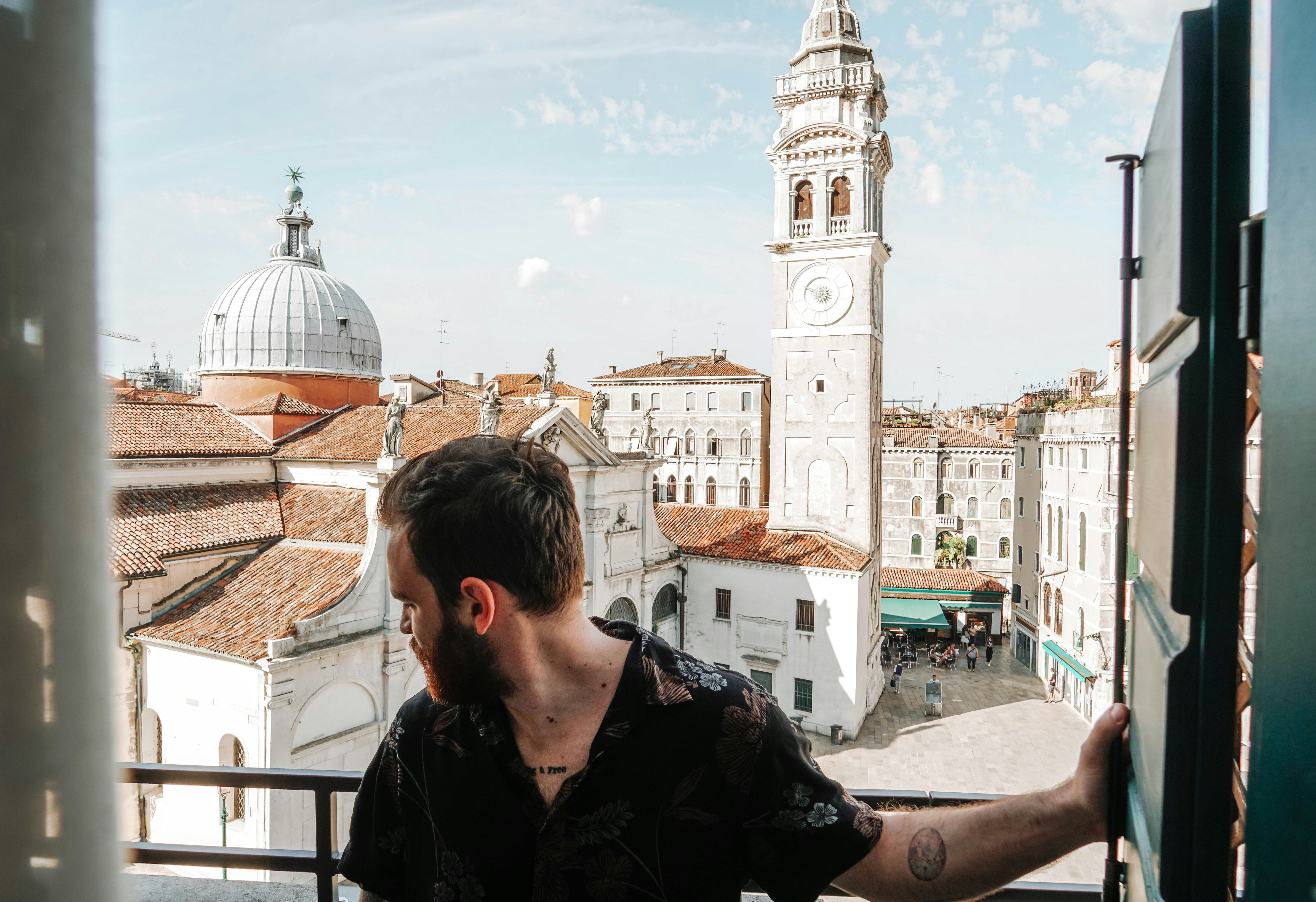 A young man gazes out from a balcony, overlooking historic architecture and a clock tower, capturing a serene moment in an urban landscape.