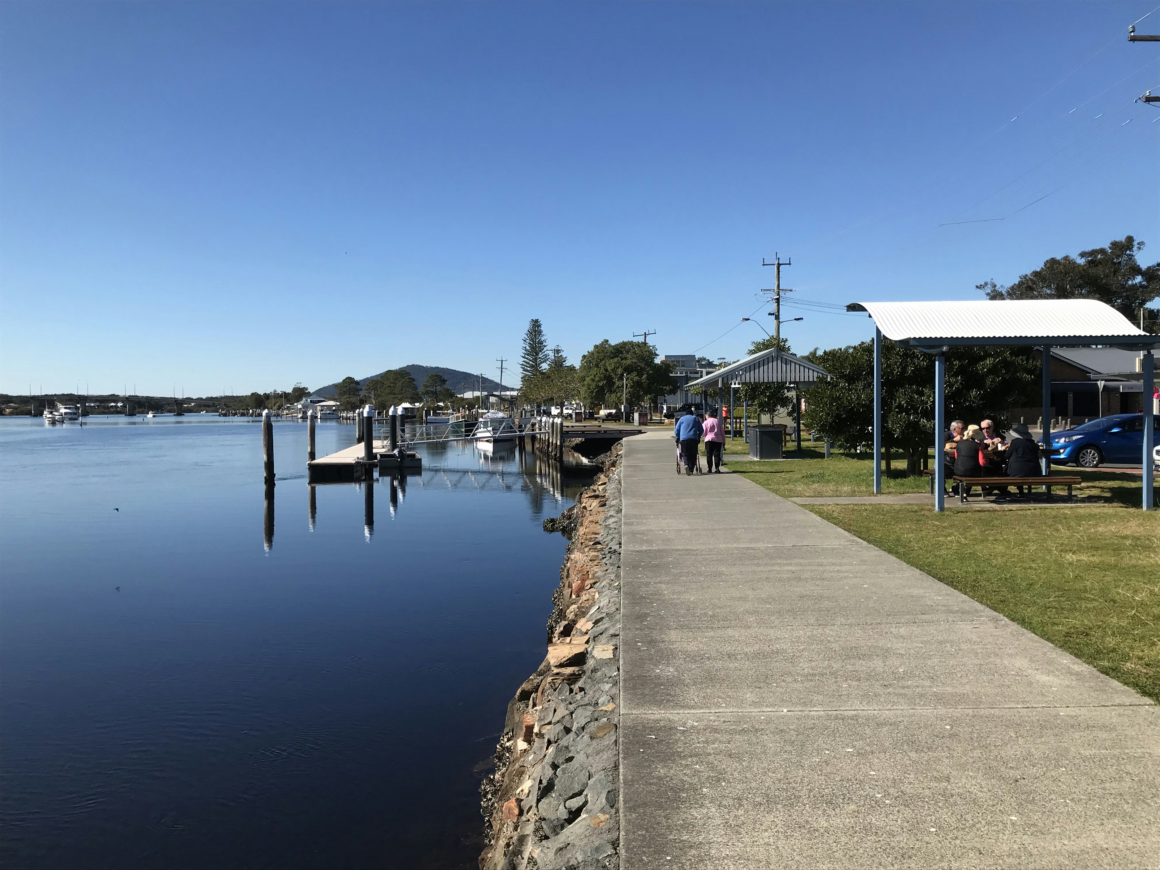 A tranquil riverside pathway lined with trees and seating areas, inviting leisurely strolls and relaxation. Boats are docked peacefully in the calm water.