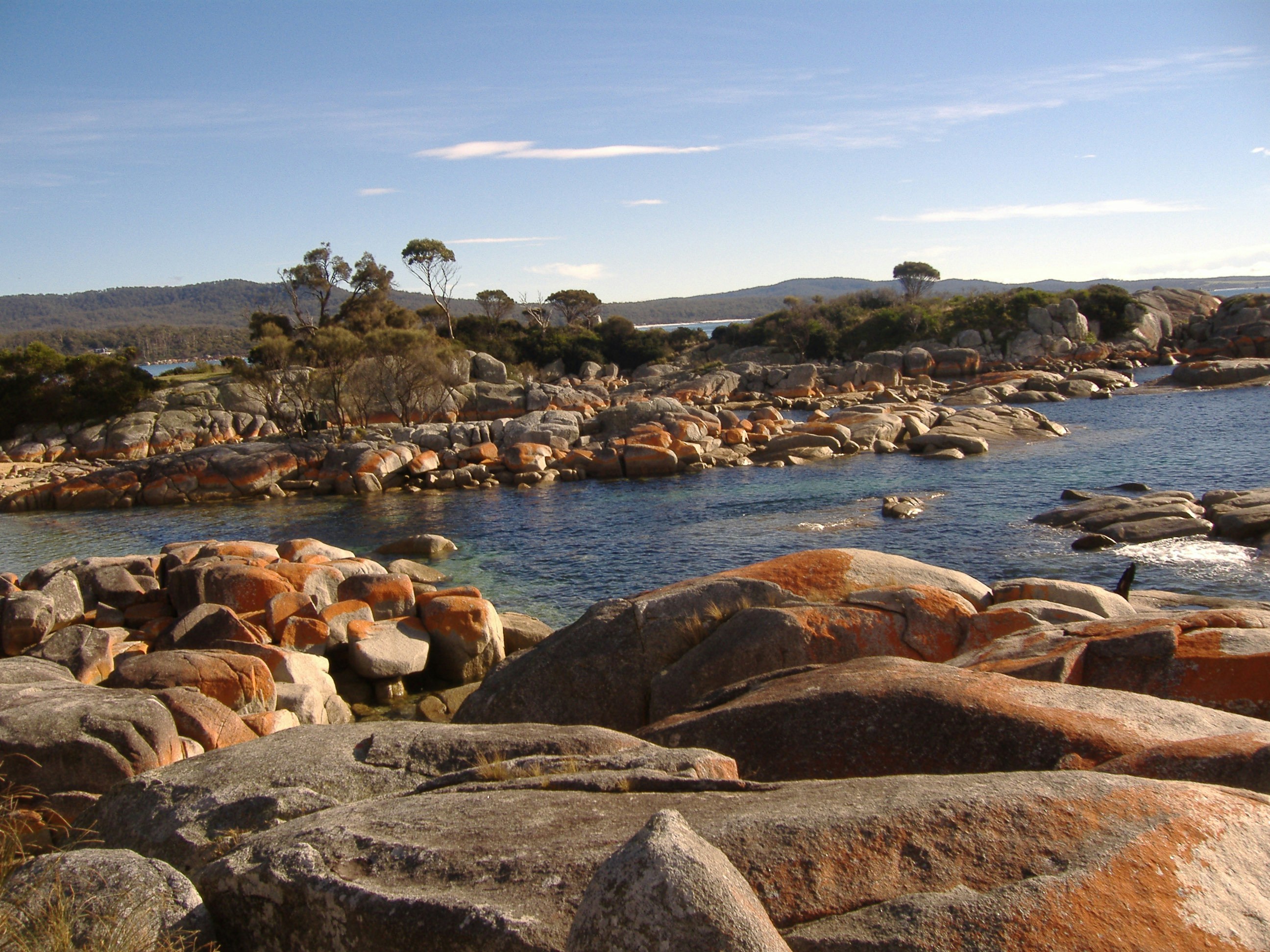 This captivating image showcases a stunning coastal landscape, featuring a tapestry of large, weathered rocks in vibrant hues of orange and grey. The serene blue water reflects the clear sky, while the distant hills and sparse trees add depth to the composition. The harmonious balance of colors and natural elements creates a tranquil and visually striking scene.