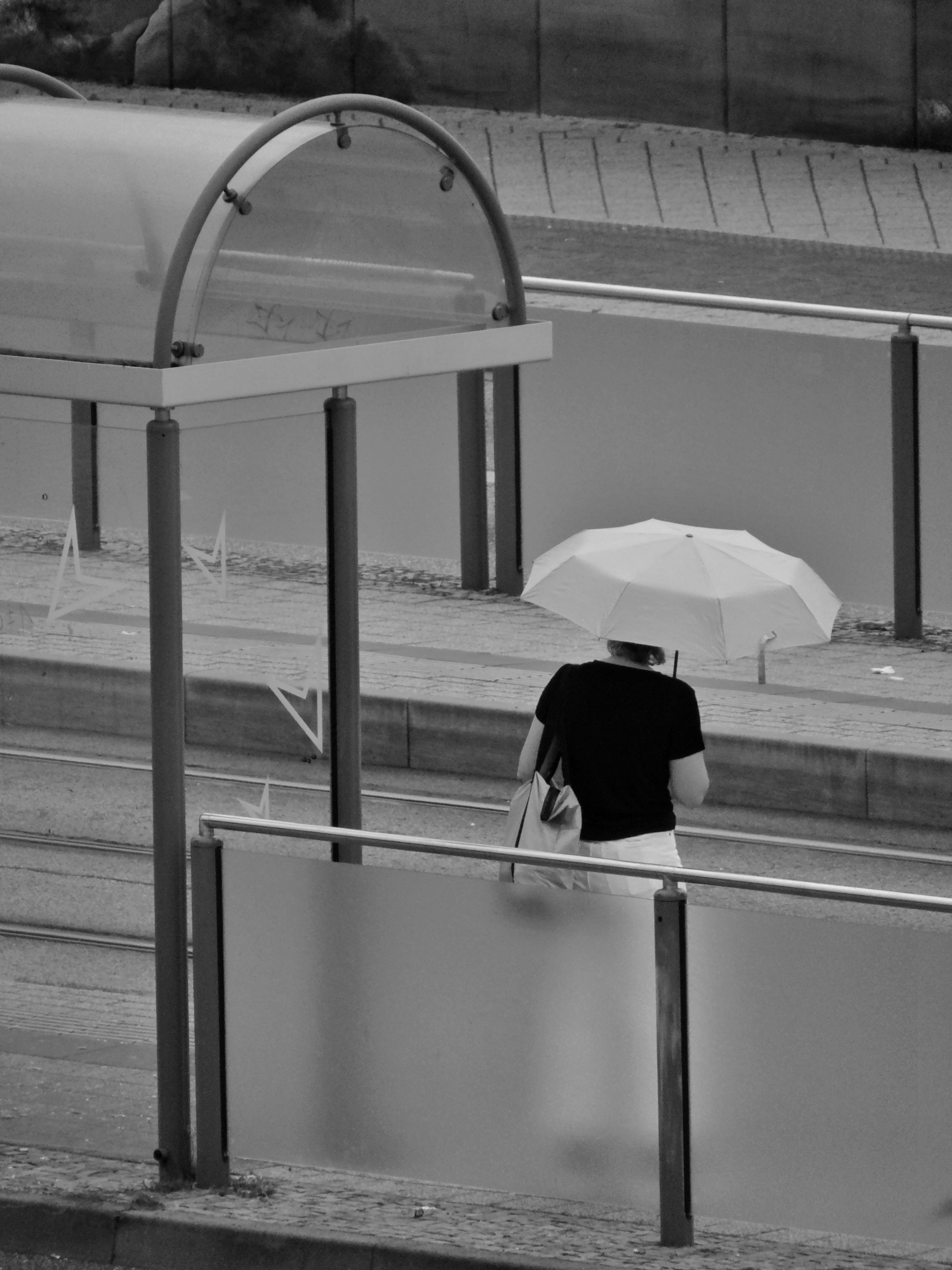 A person stands at a bus stop holding a white umbrella, dressed in black and white, surrounded by a minimalist urban backdrop.