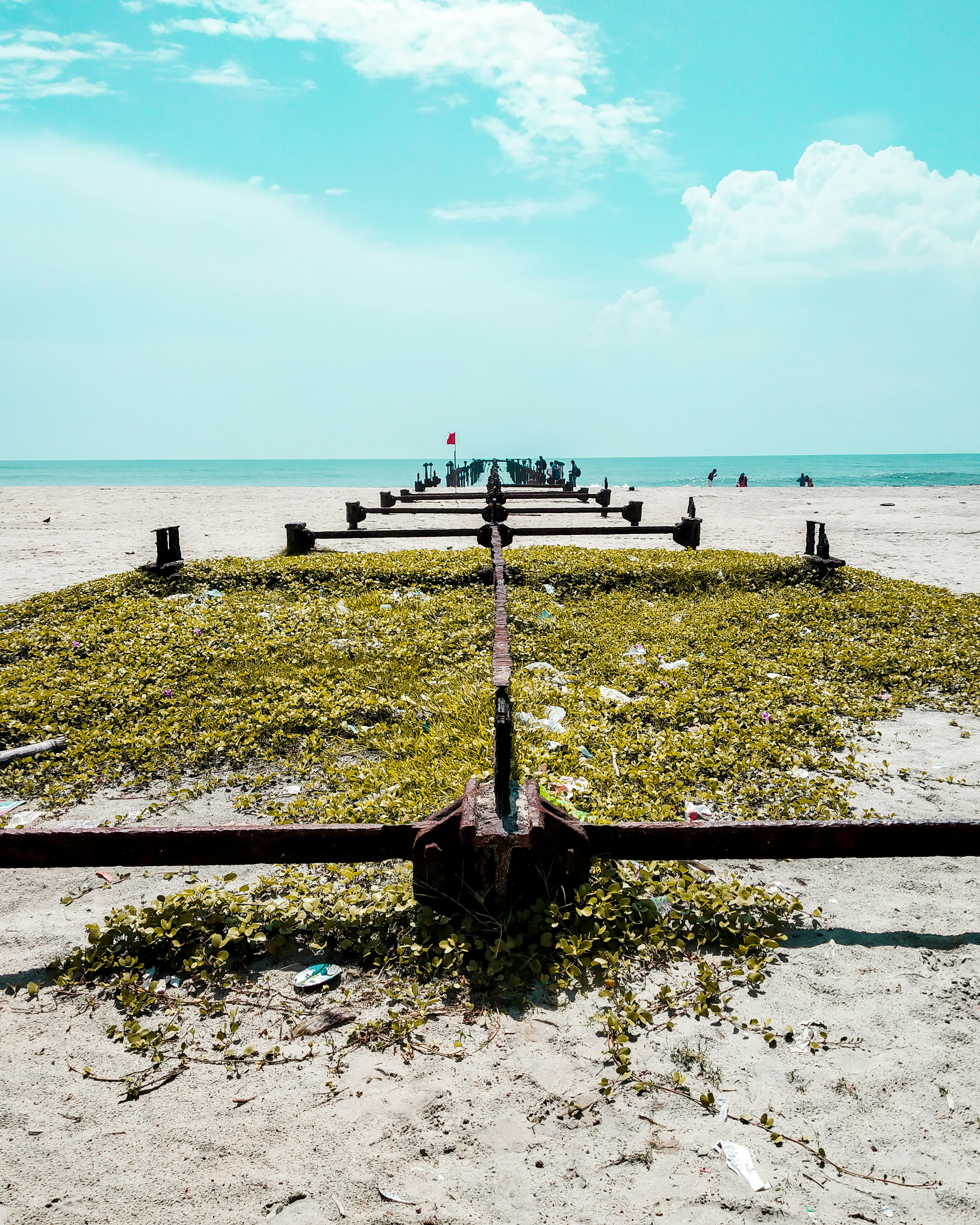 Rusty metal beams covered in seaweed stretch toward the horizon on a sandy beach, with distant figures enjoying the seaside. A vibrant blue sky adds depth to the scene.