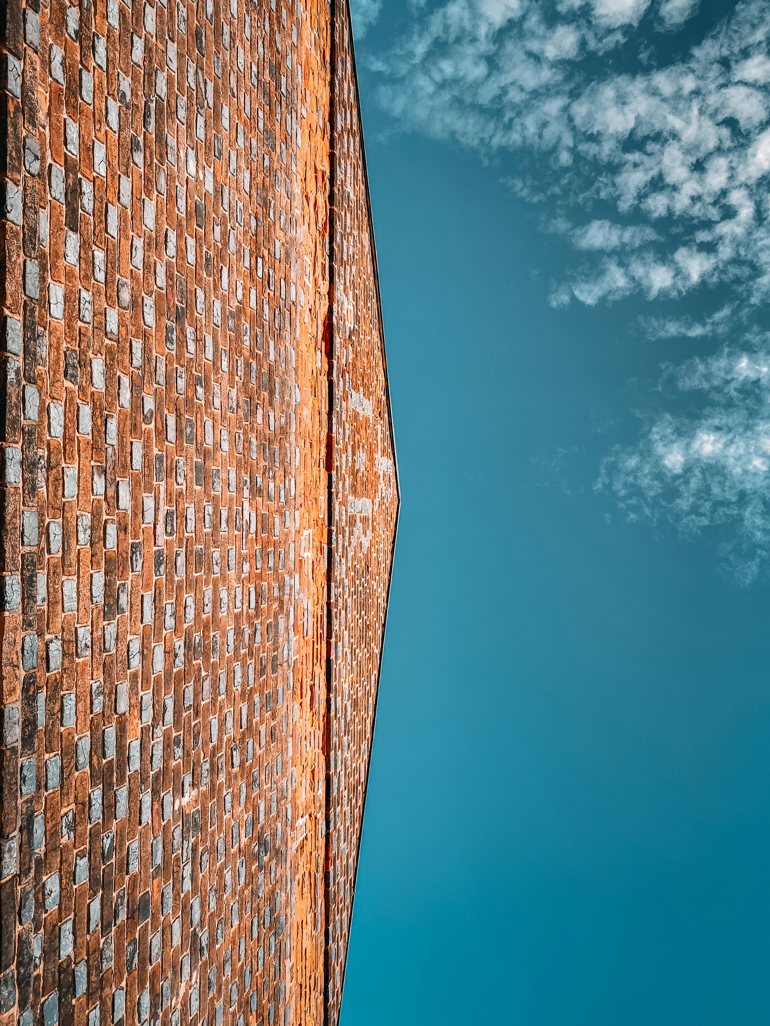 Close-up of a textured brick wall ascending towards a clear blue sky with wispy clouds.