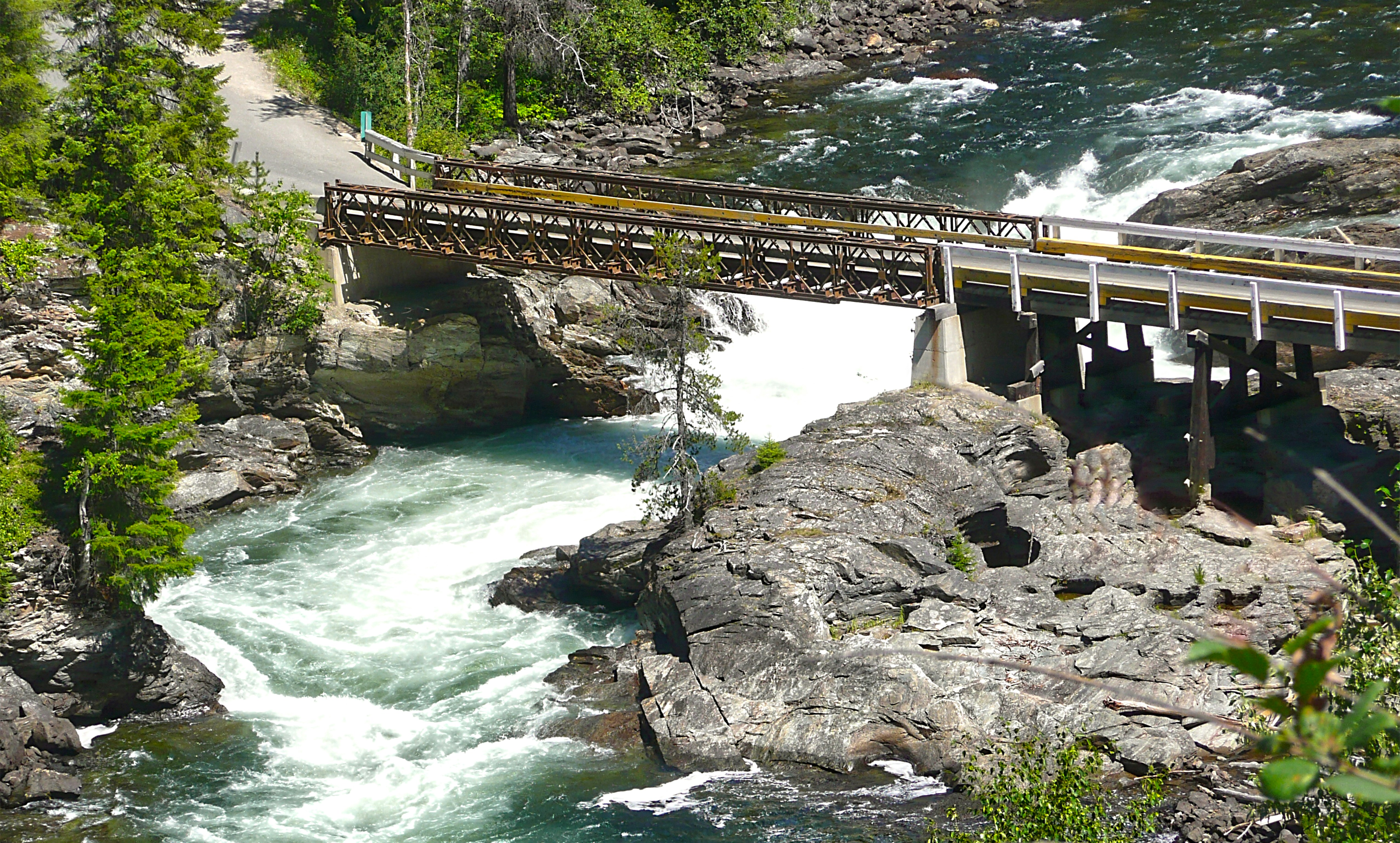 A rustic wooden bridge spans a rushing river, surrounded by lush greenery and rocky terrain. The dynamic flow of water contrasts with the stability of the bridge.