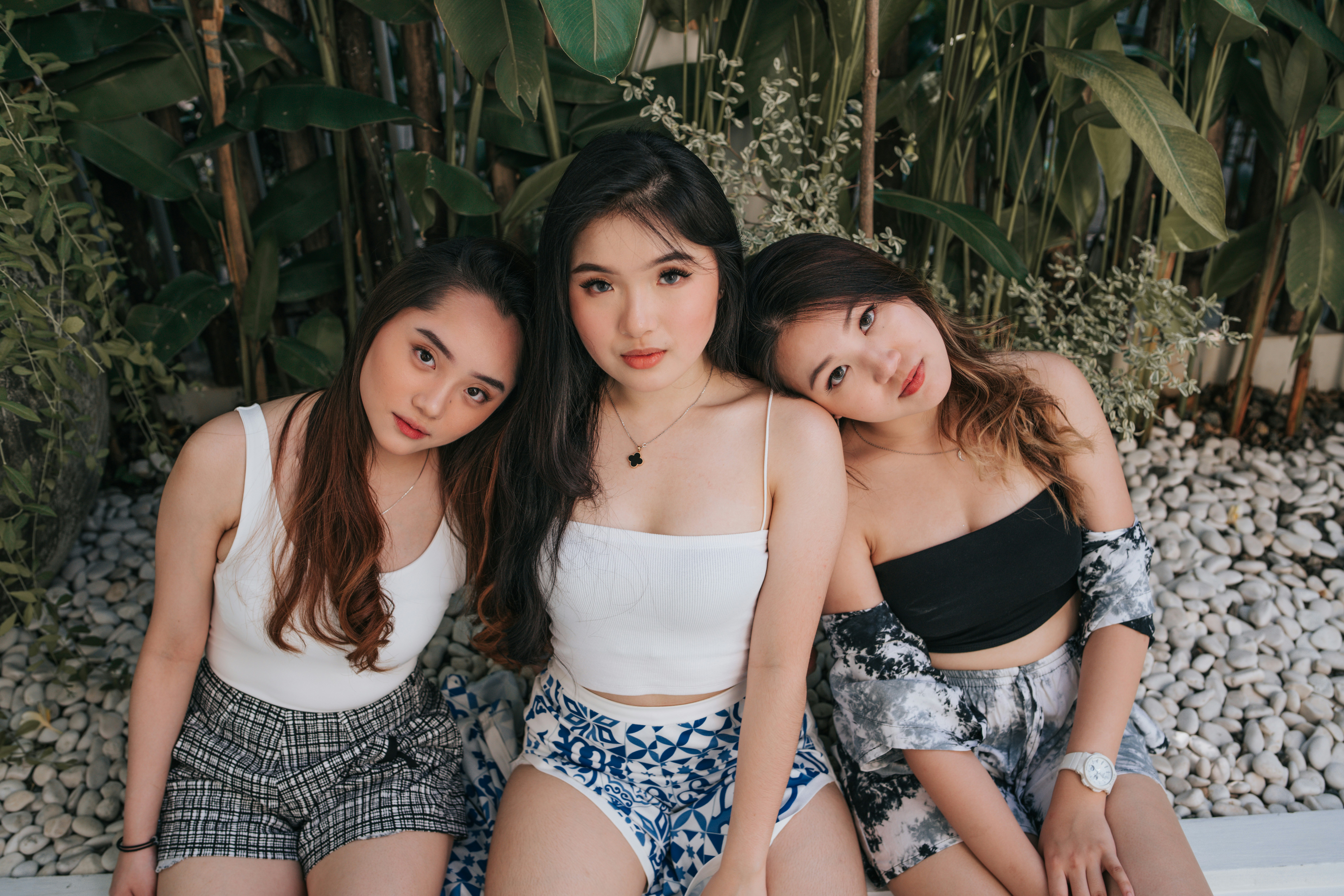 Three young women seated together against a backdrop of lush greenery, showcasing their stylish summer outfits and joyful expressions.