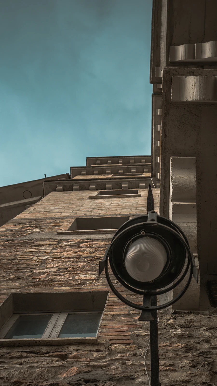 Low-angle view of a tall brick building against a blue sky, with a round industrial lamp in the foreground. The strong vertical lines and textured masonry create a stark urban composition.