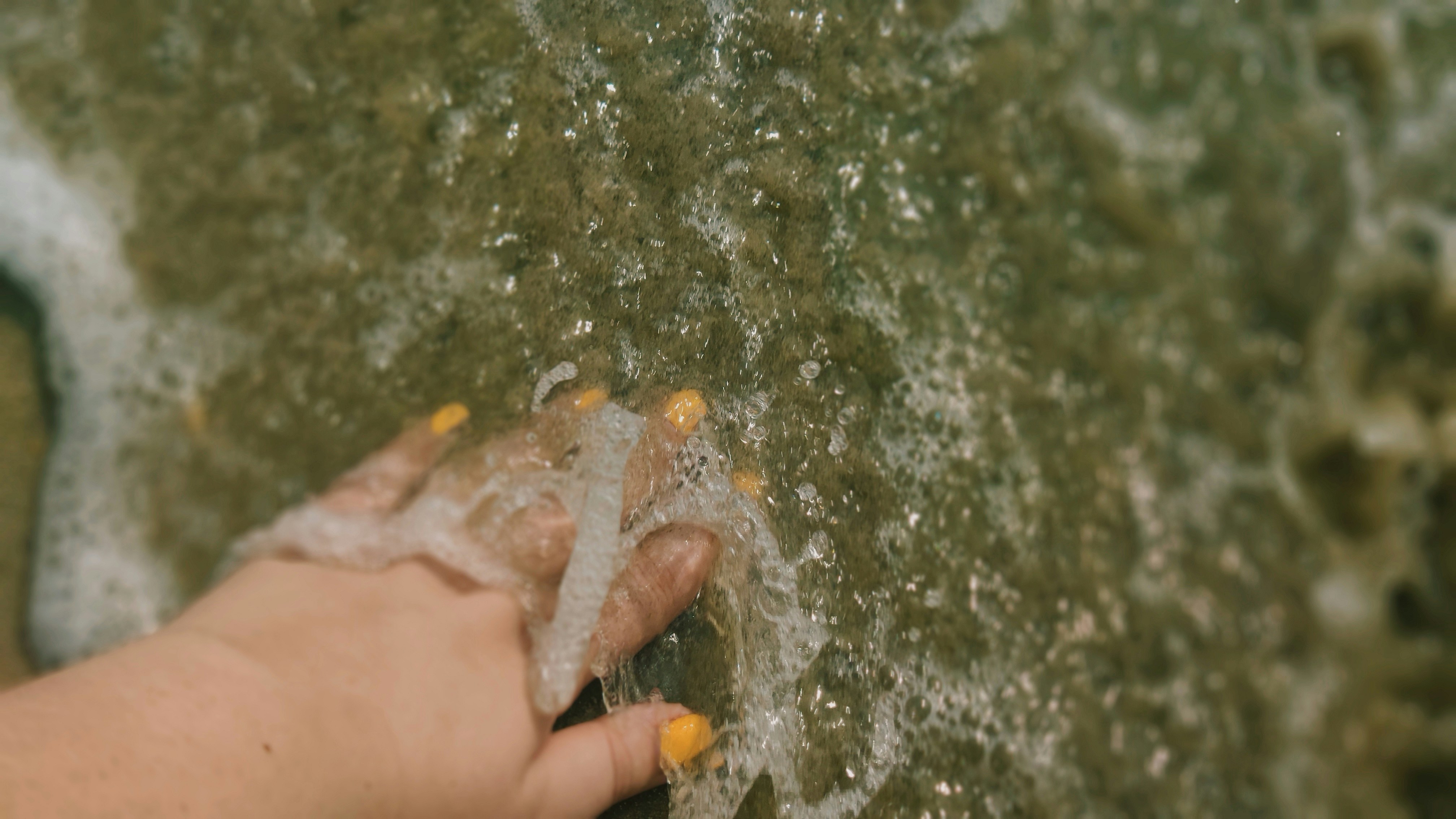 Close-up of a hand with yellow nails touching a mossy, wet rock, highlighting textured surfaces and droplets. The scene emphasizes natural texture and tactile contrast.
