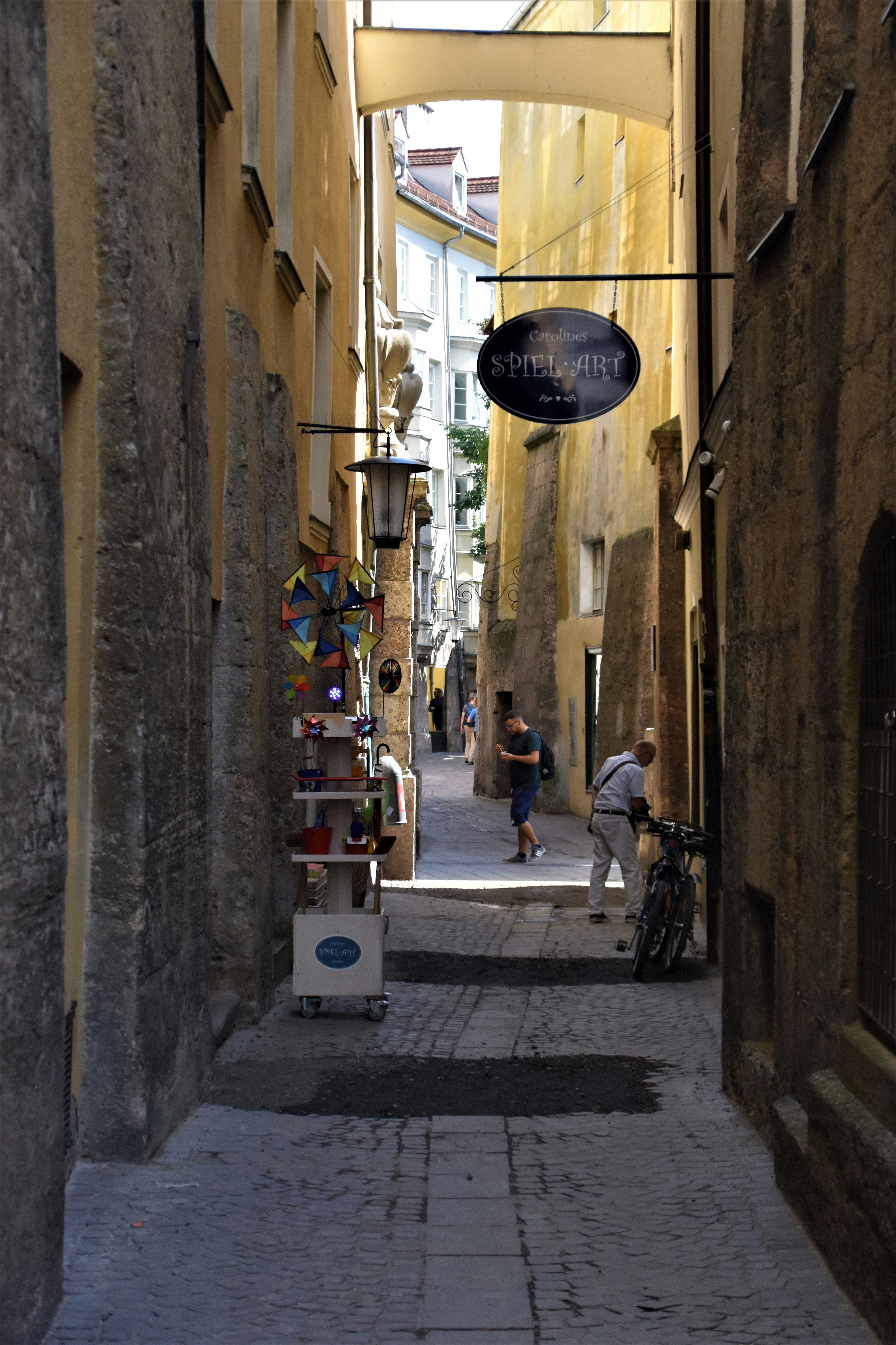 Narrow cobblestone alleyway lined with charming buildings, featuring a colorful display of pinwheels and a bicycle parked against the wall. A pedestrian strolls through the sunlit path.