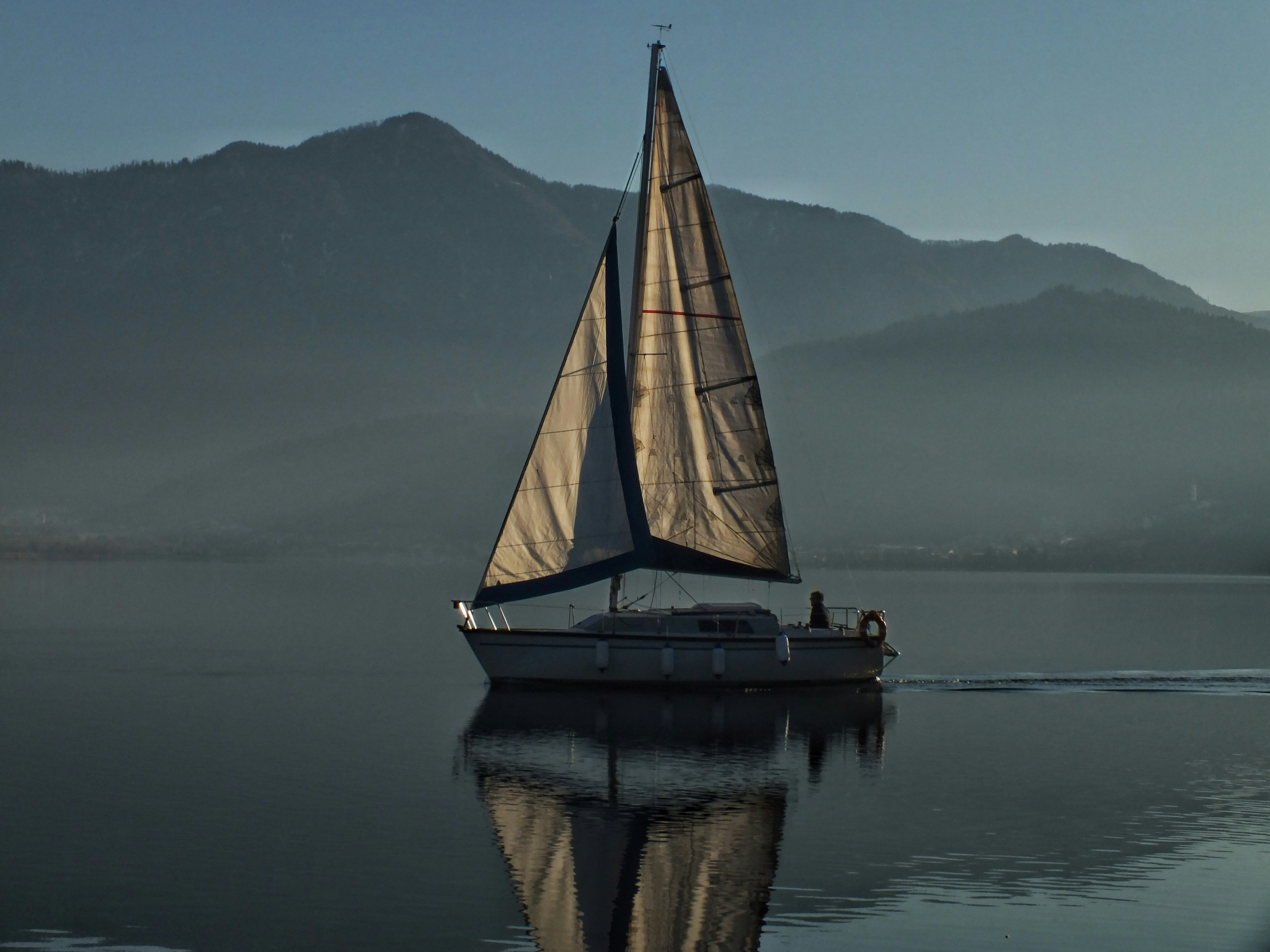 Sailboat gliding across a calm lake, surrounded by misty mountains at dawn. The reflection in the water enhances the tranquil atmosphere.