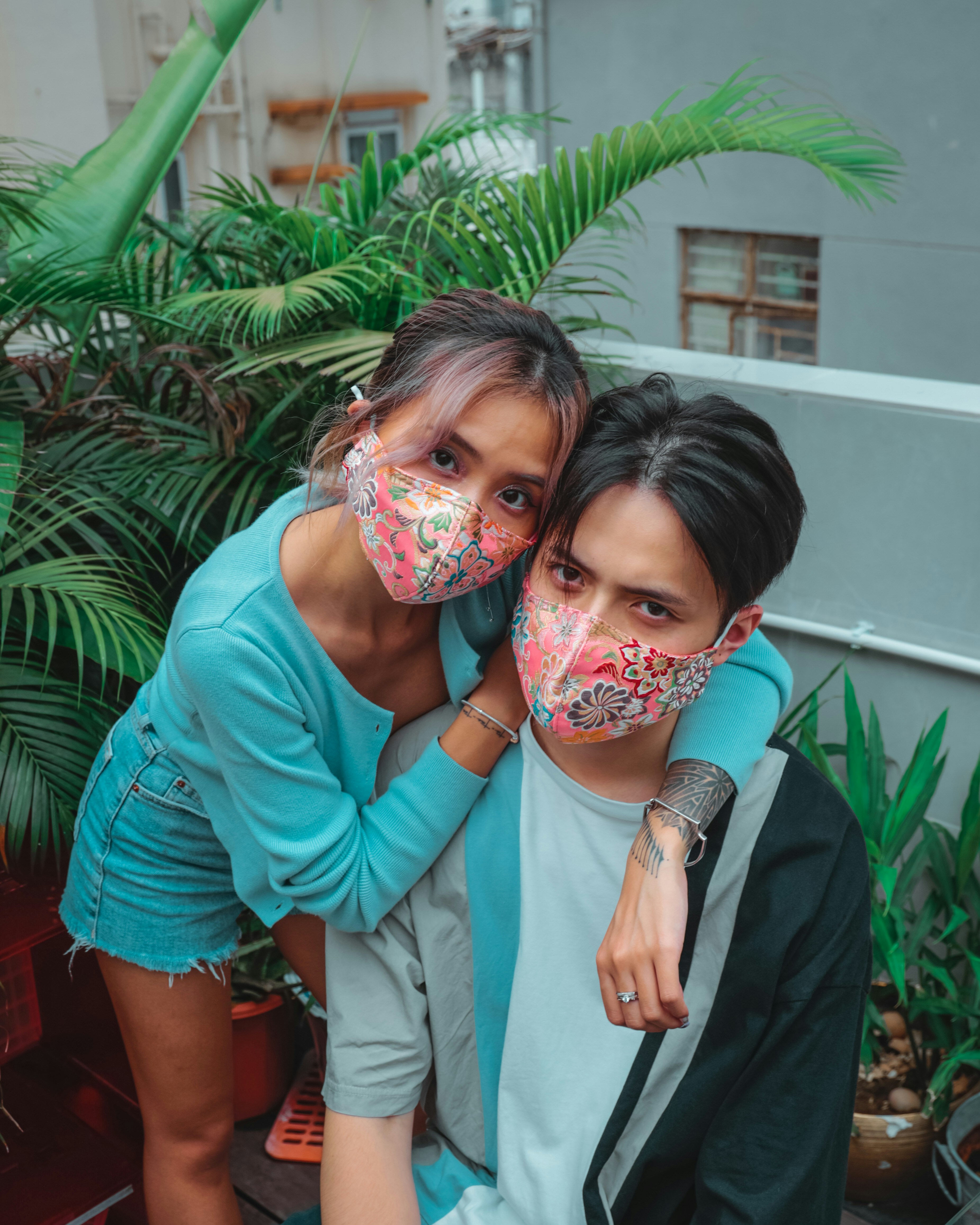 Couple in vibrant floral masks and teal clothing, surrounded by lush greenery on a rooftop.