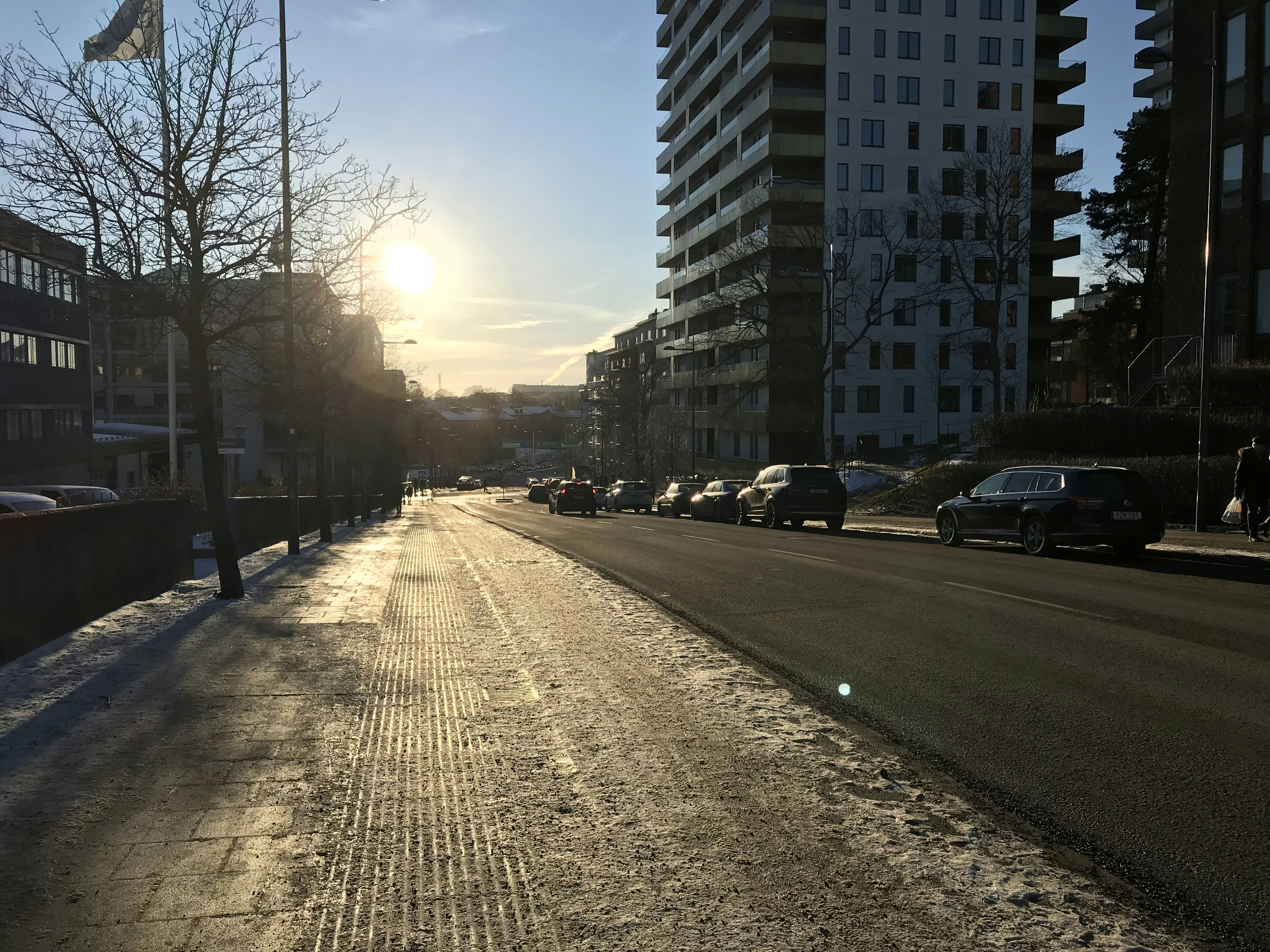 Sunset casts a warm glow over a snowy street lined with modern buildings and parked cars.