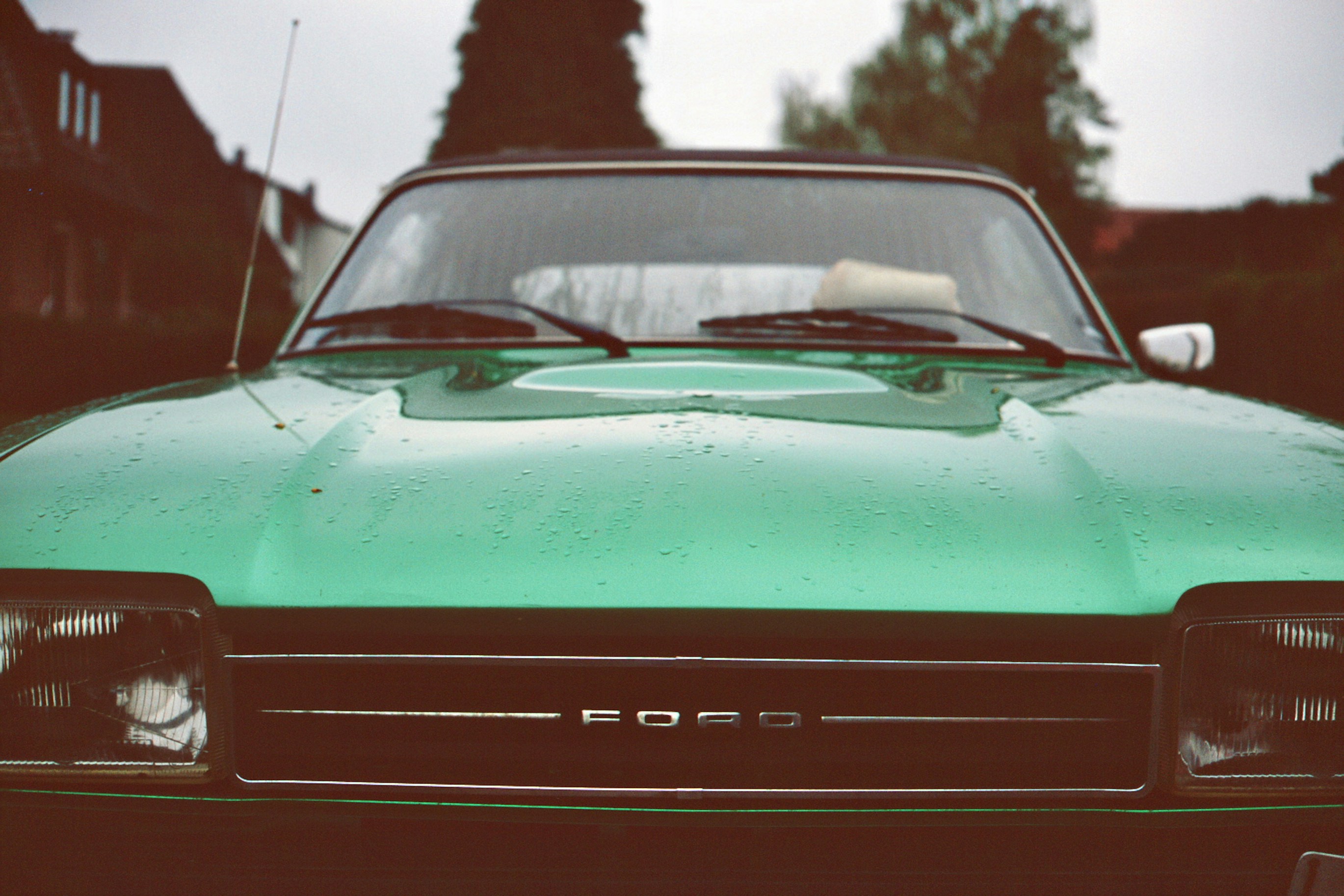 Close-up of a vintage Ford front with a glossy emerald hood, rain droplets, and chrome grille, photographed head-on.