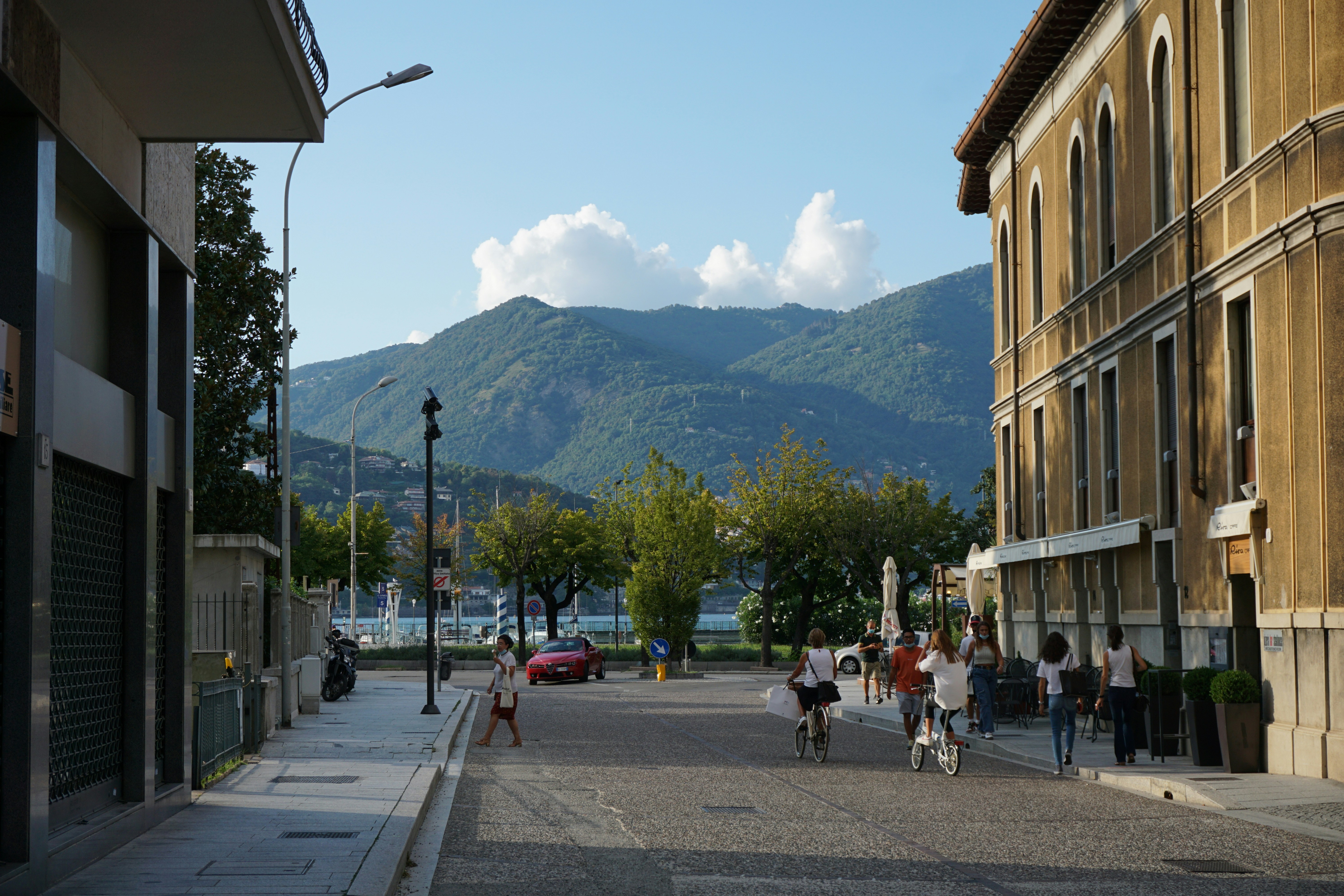 Bicyclists and pedestrians enjoy a leisurely afternoon along a lakeside promenade, framed by mountains and vibrant greenery.