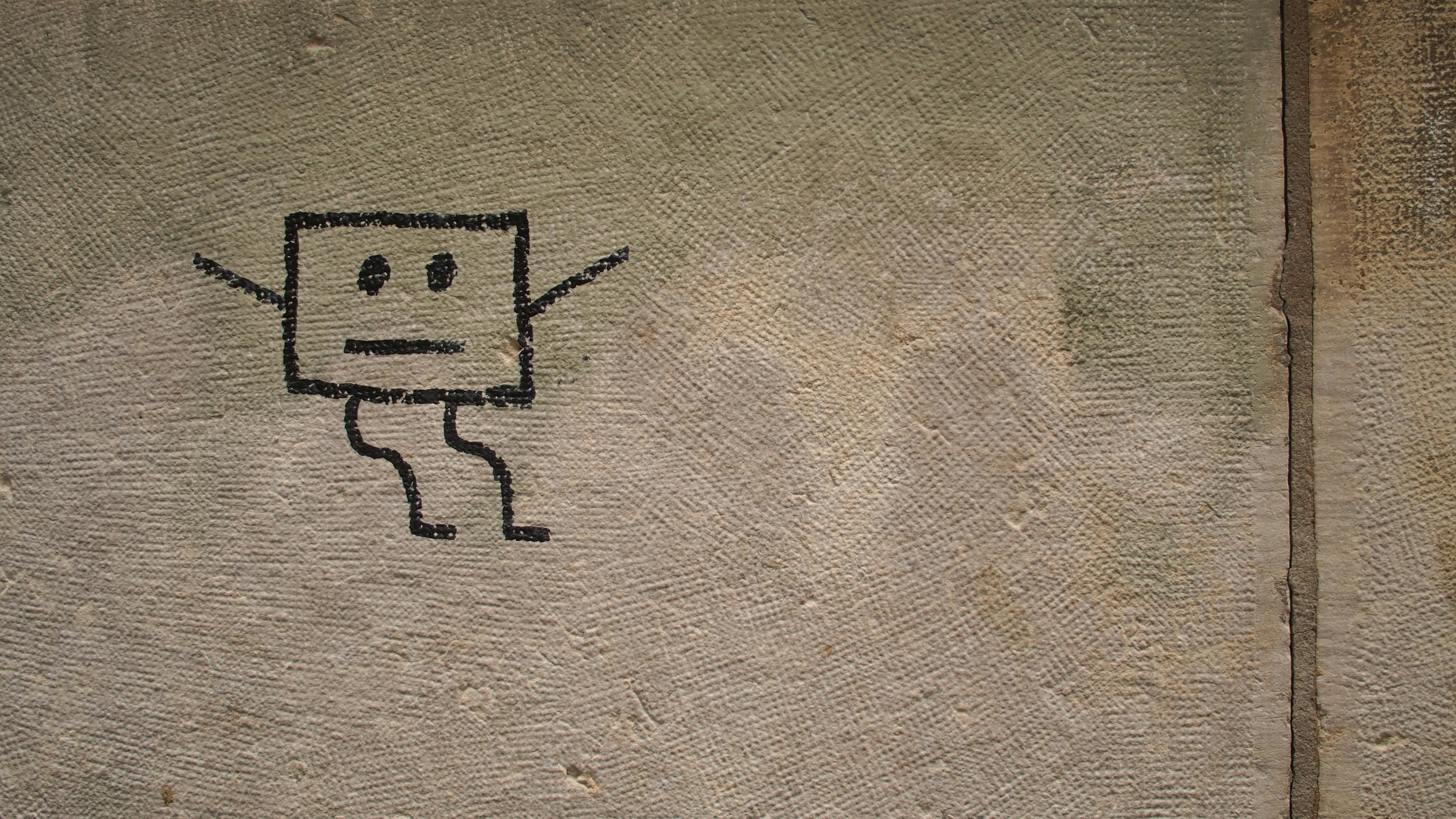 Simple cartoon character drawn on a textured wall, showcasing playful street art. The character features a rectangular head and stick-like limbs.