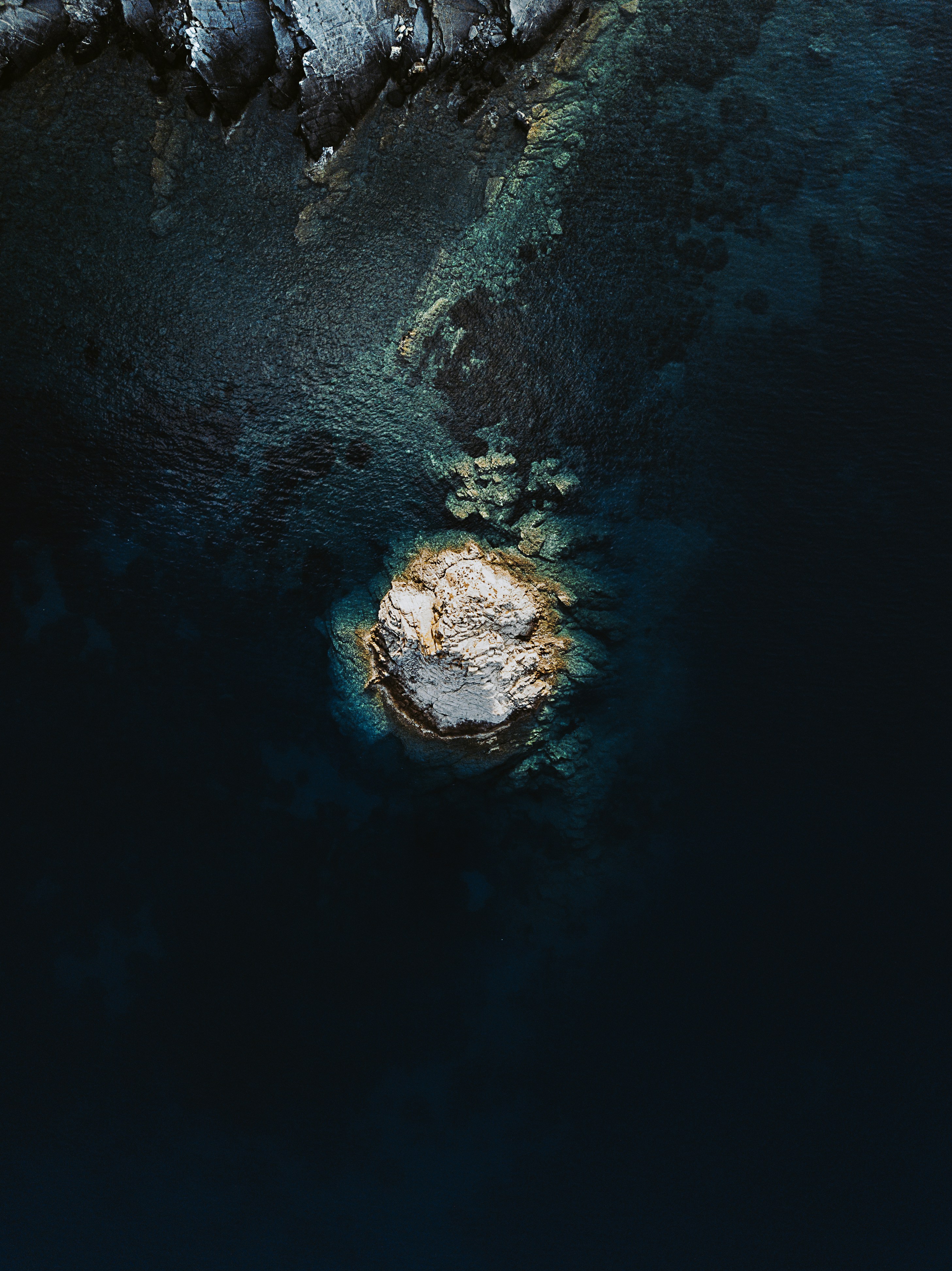 Aerial view of a solitary rock formation surrounded by deep blue waters, showcasing the contrast between land and sea. The scene highlights the natural beauty of the coastline.