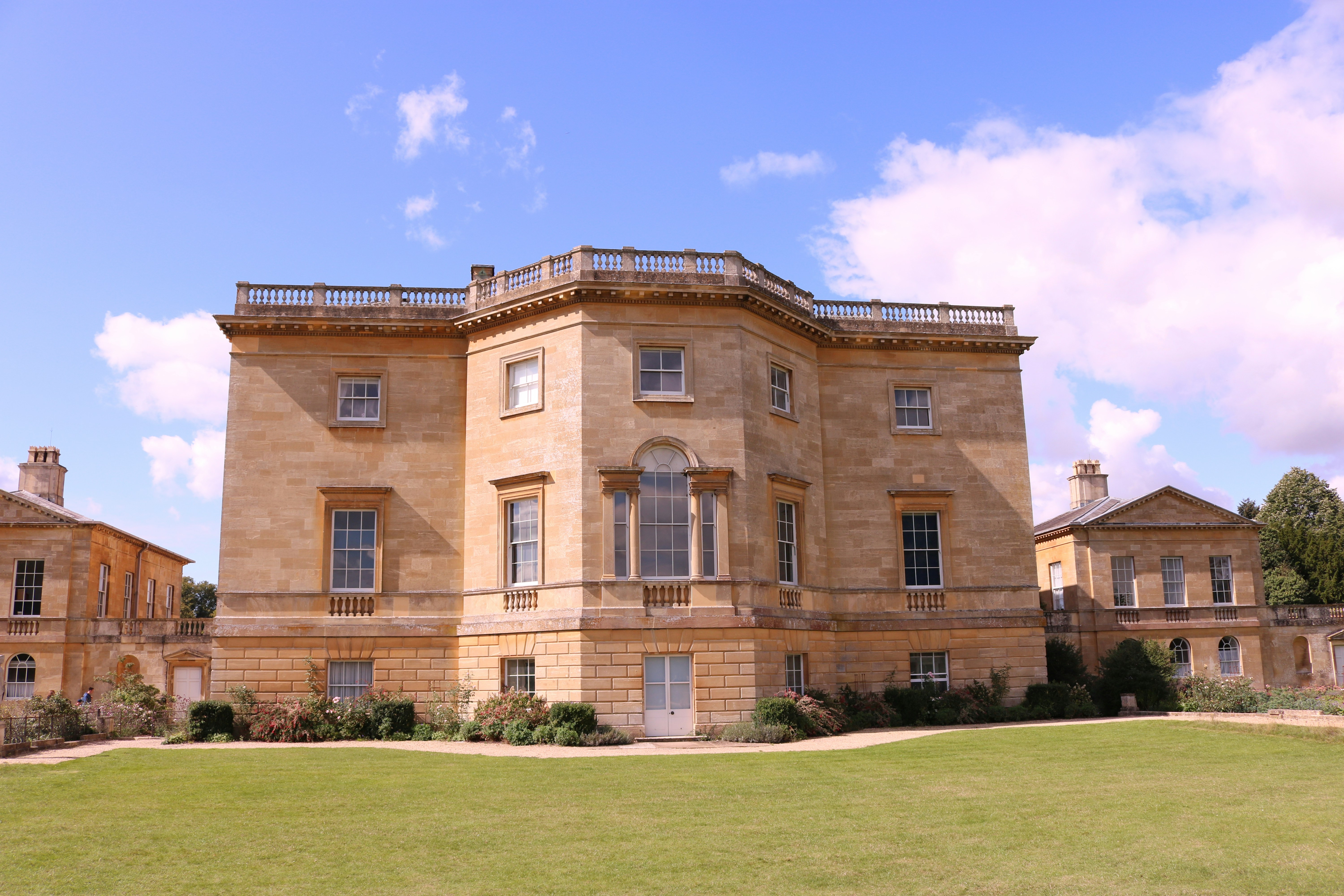 Historic stone mansion with symmetrical architecture set against a bright blue sky and fluffy clouds.