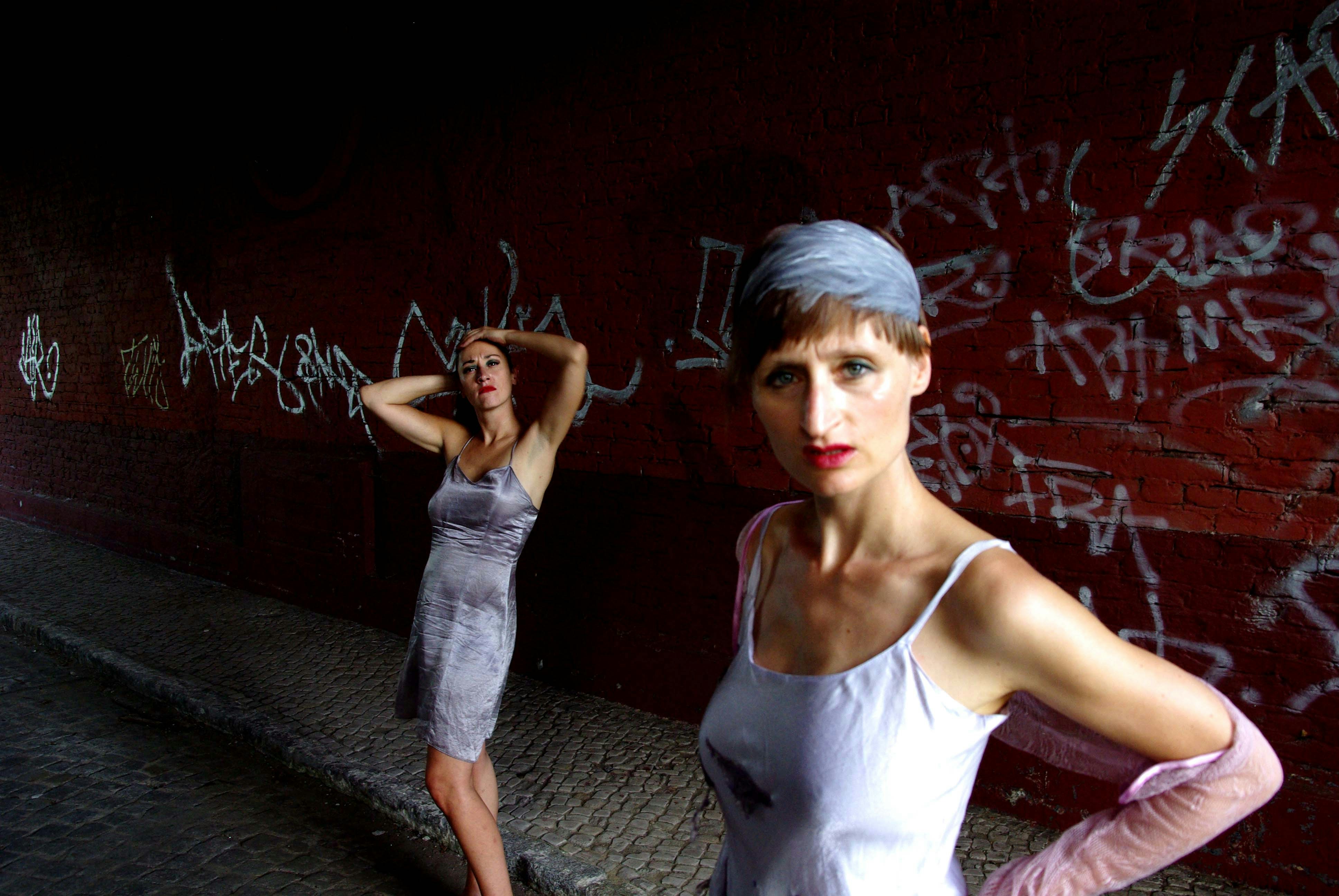 Two women posing in a graffiti-covered alley wearing satin dresses. The foreground figure with blue hair dominates the frame, creating a bold urban portrait.