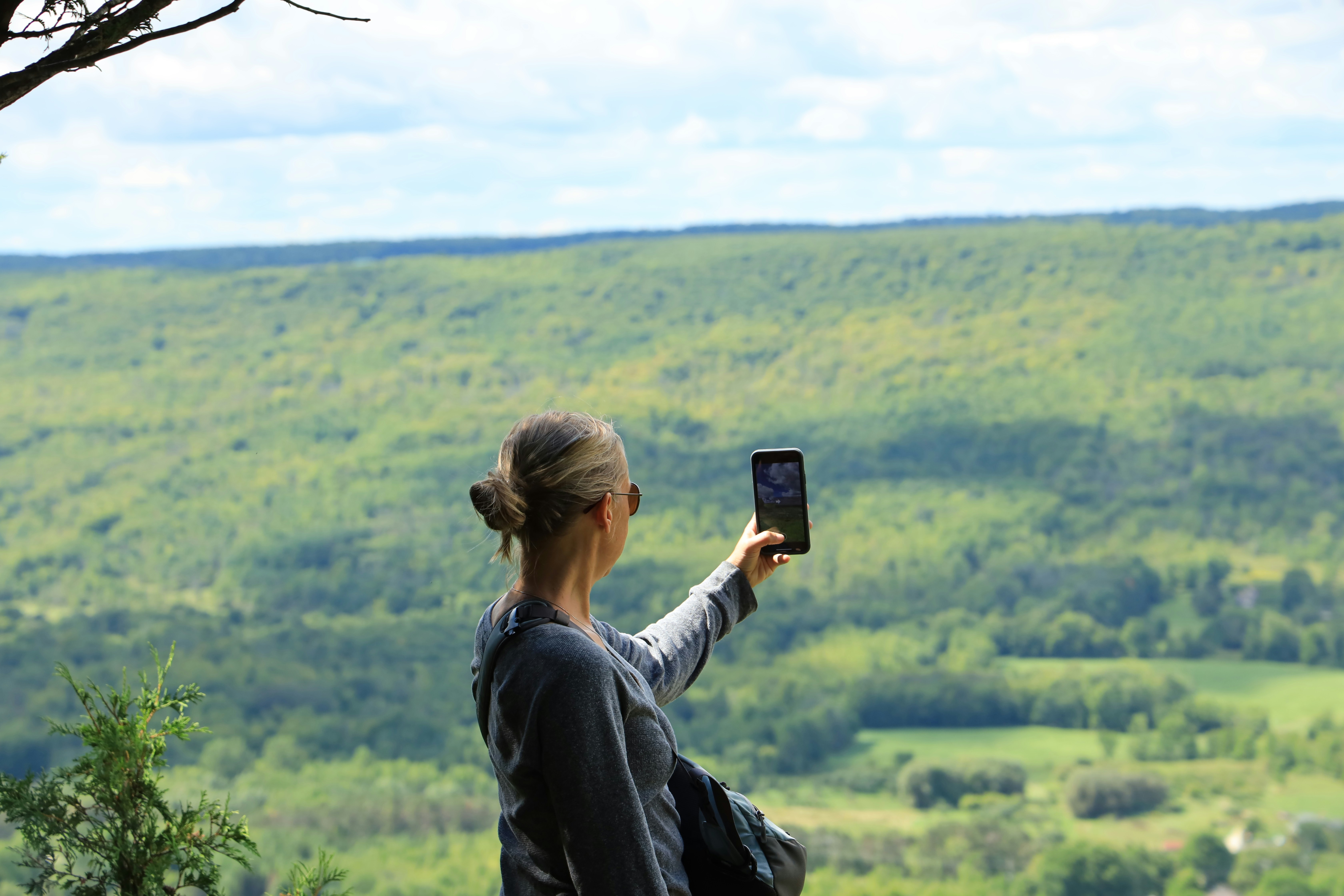 Person taking a selfie with a smartphone against a lush green landscape. The scene showcases the beauty of nature in the background.