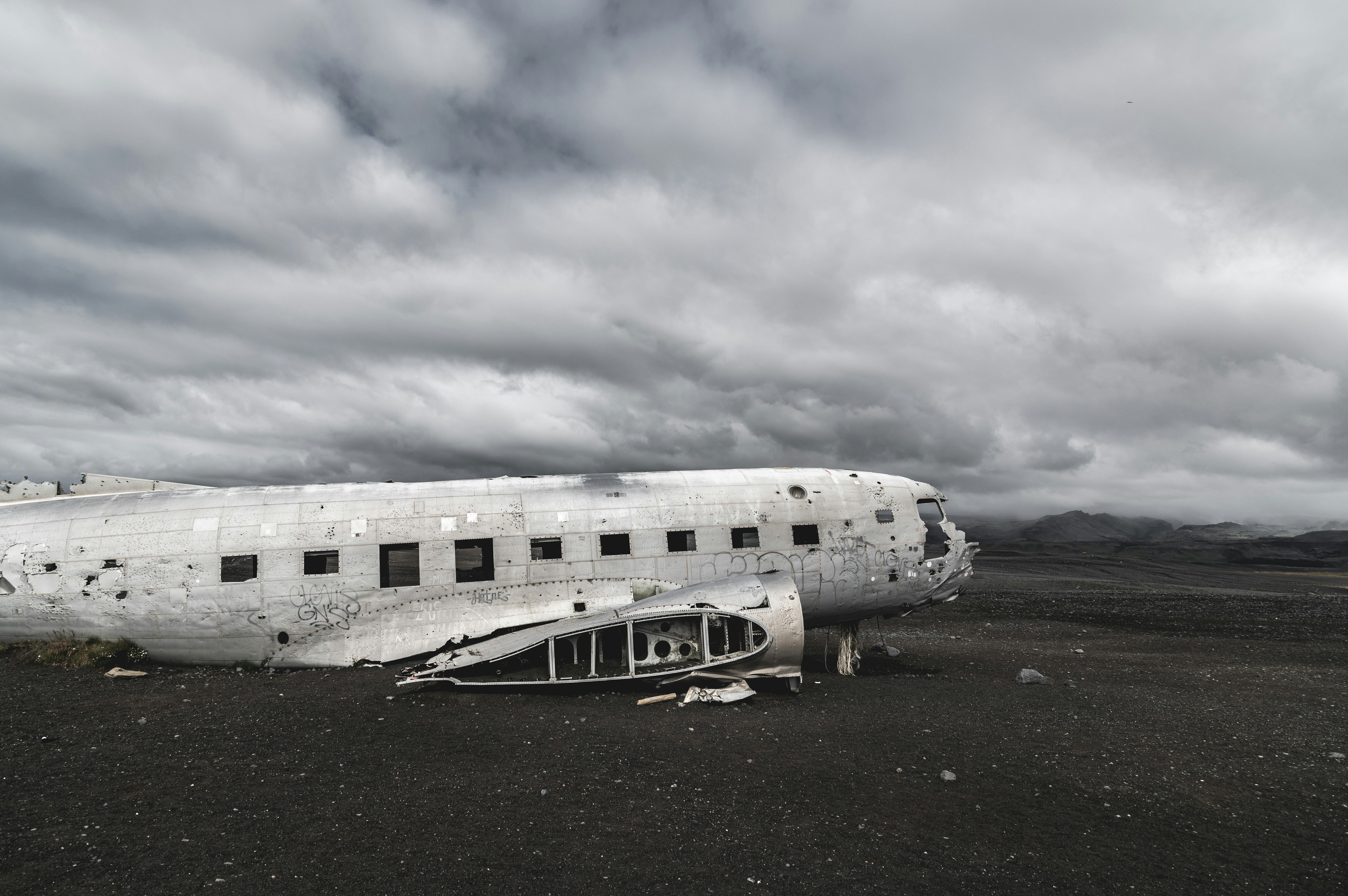 Abandoned airplane wreckage rests on barren volcanic sand under dramatic cloudy skies.