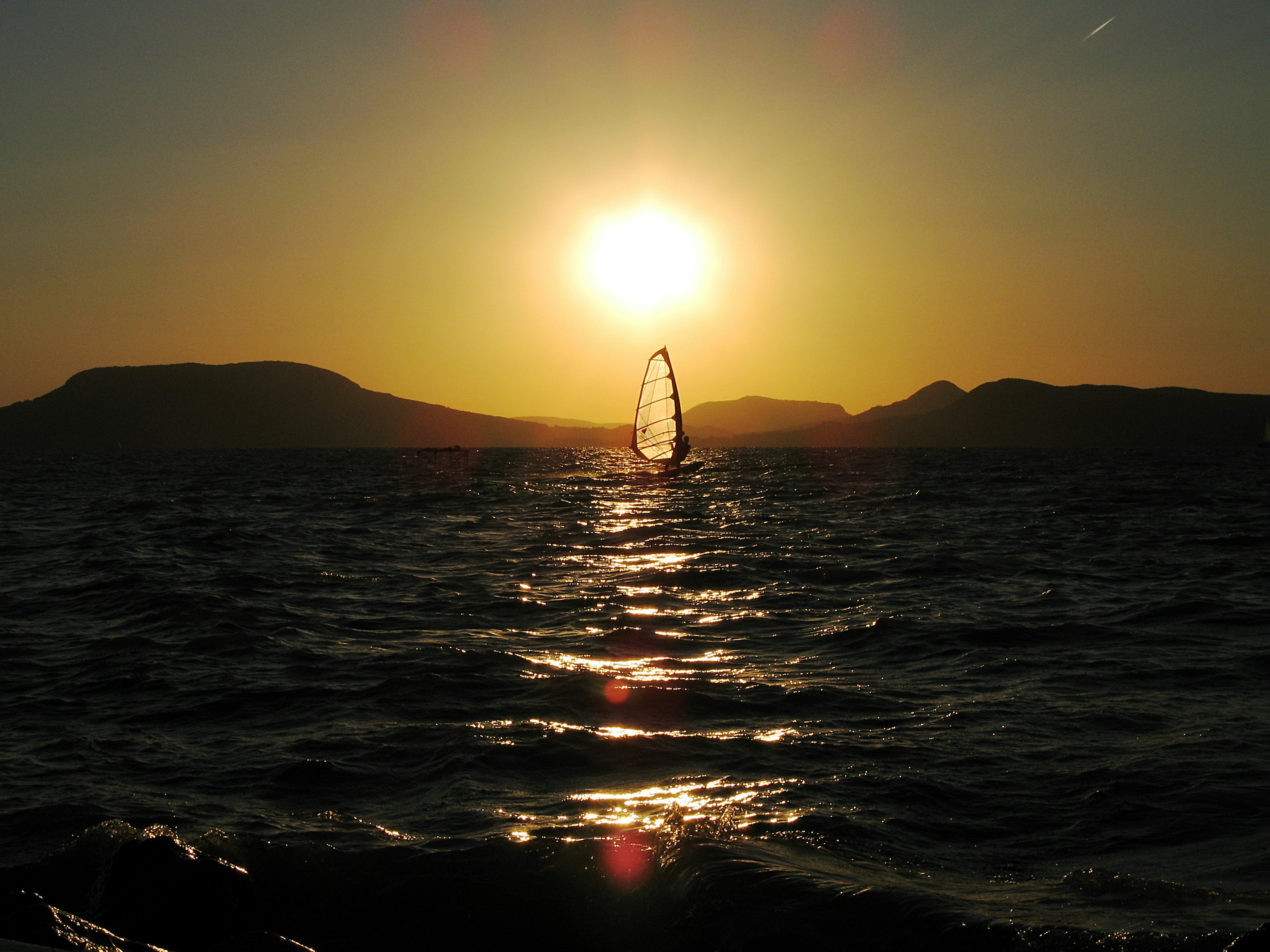 Evening photograph of a windsurfer silhouetted against a golden sunset over a shimmering sea.