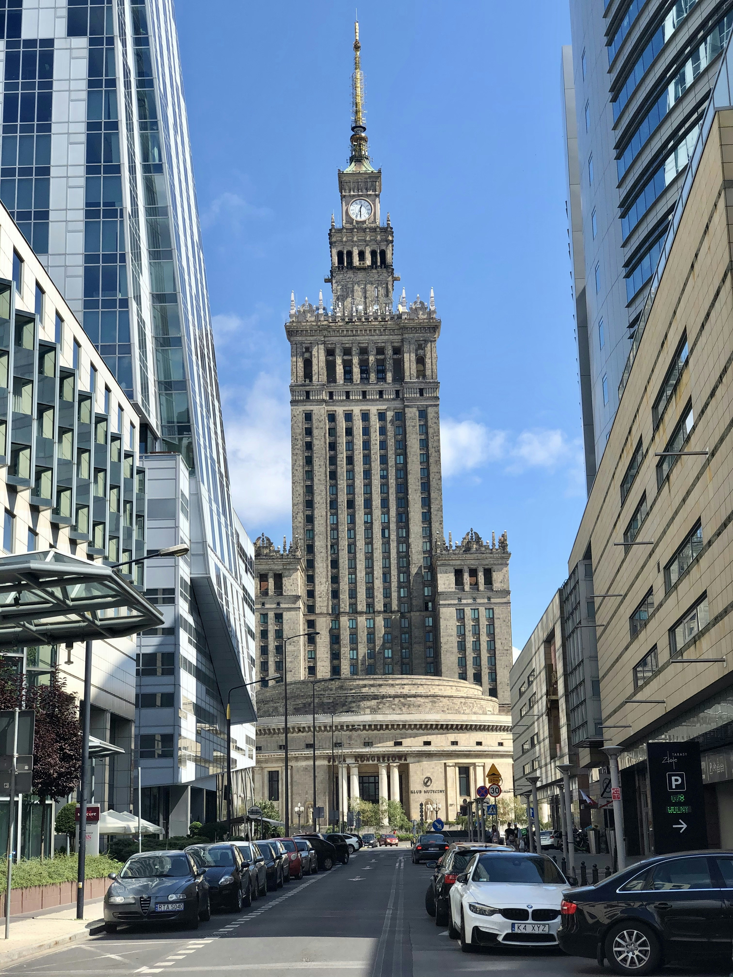 The Palace of Culture and Science towers over a bustling city street, framed by sleek modern buildings and parked cars. A clear blue sky enhances the scene.