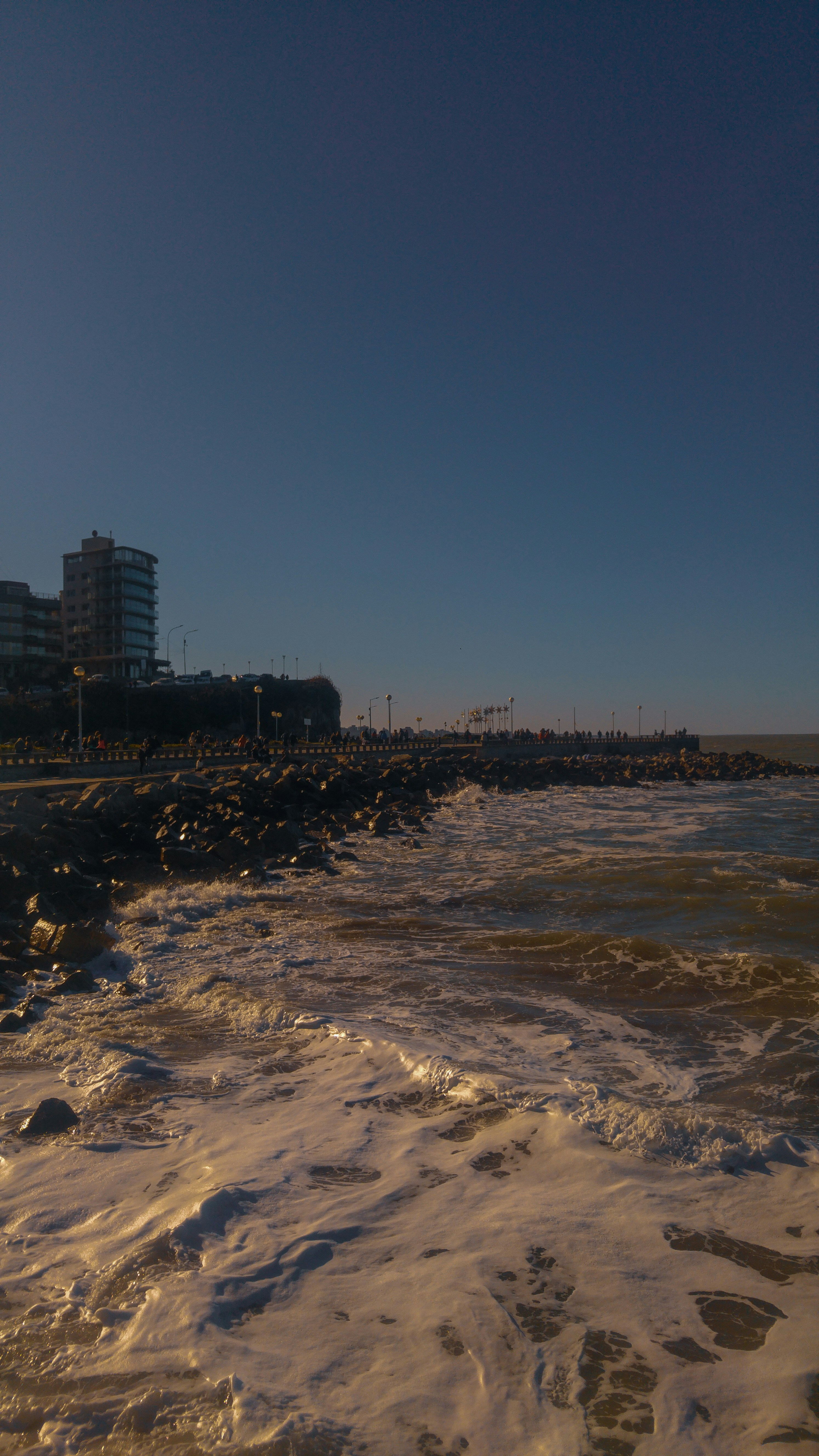 Waves crashing against rocky shore under a clear blue sky, with distant buildings lining the coast. The scene captures the serene essence of a coastal evening.