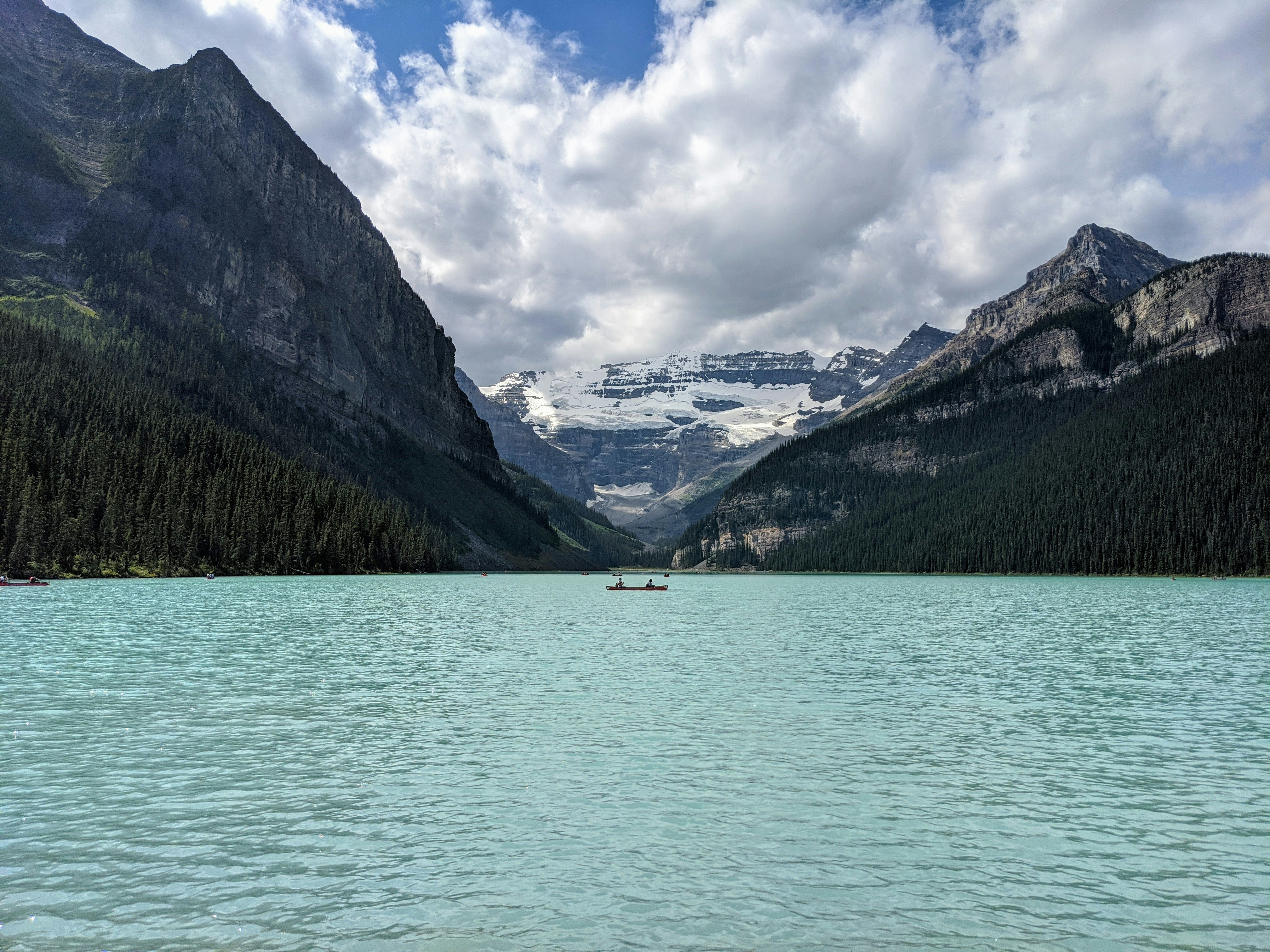Canoe gliding on a turquoise lake surrounded by towering mountain peaks under a partly cloudy sky.