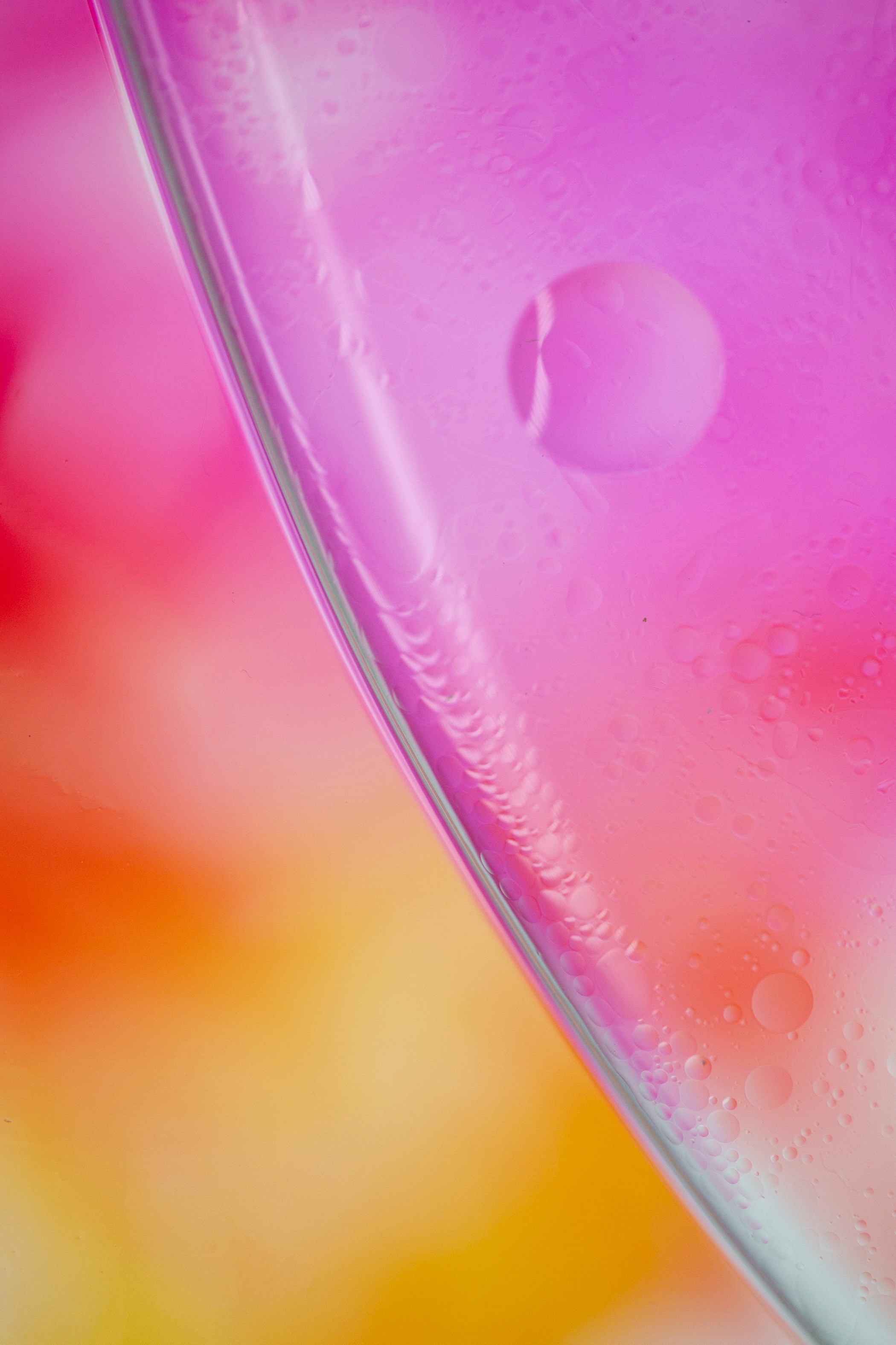 Close-up of a glass with vibrant pink liquid, featuring bubbles and droplets against a blurred, colorful background.