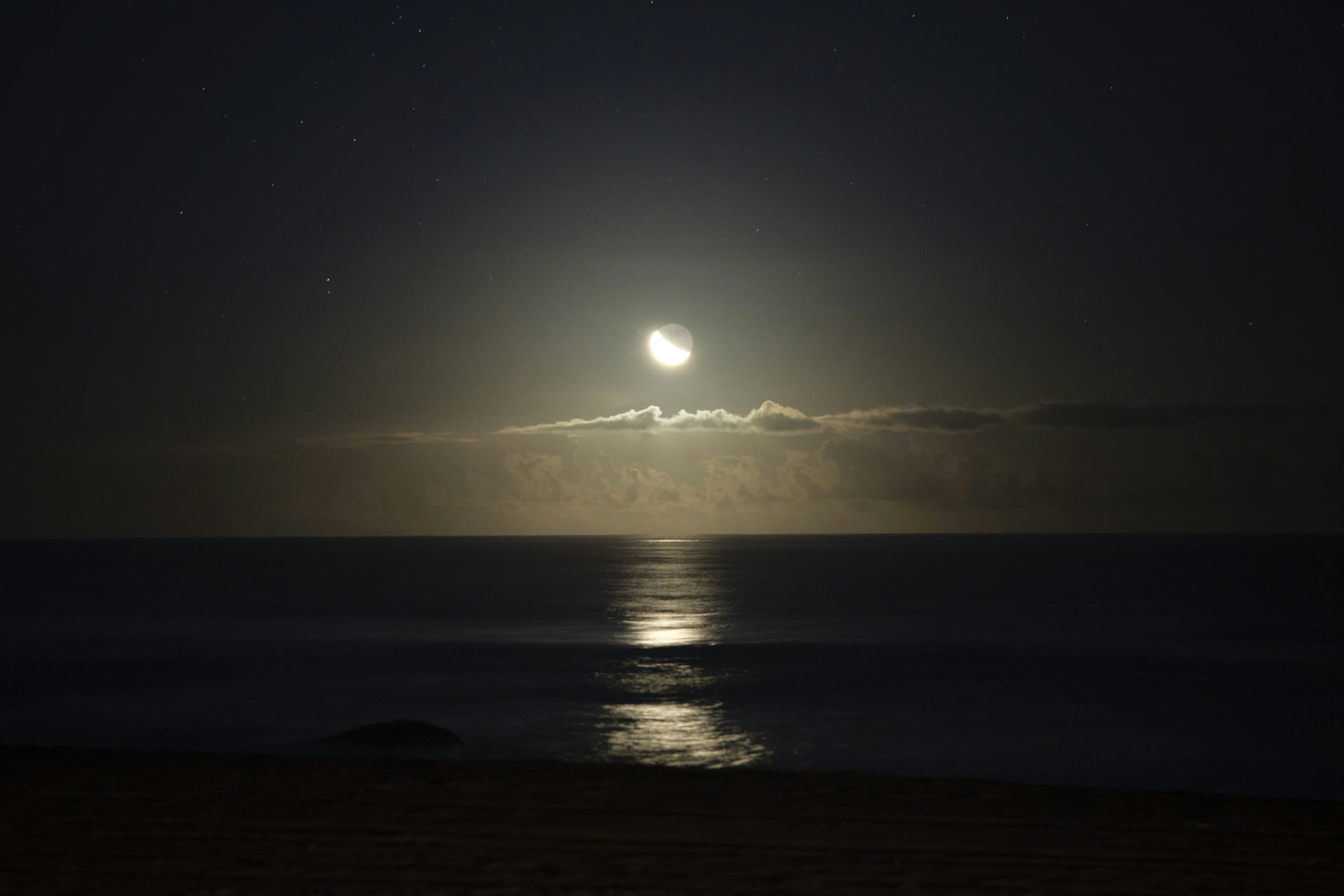 Moonlight reflecting on the calm ocean at night under a partially cloudy sky.
