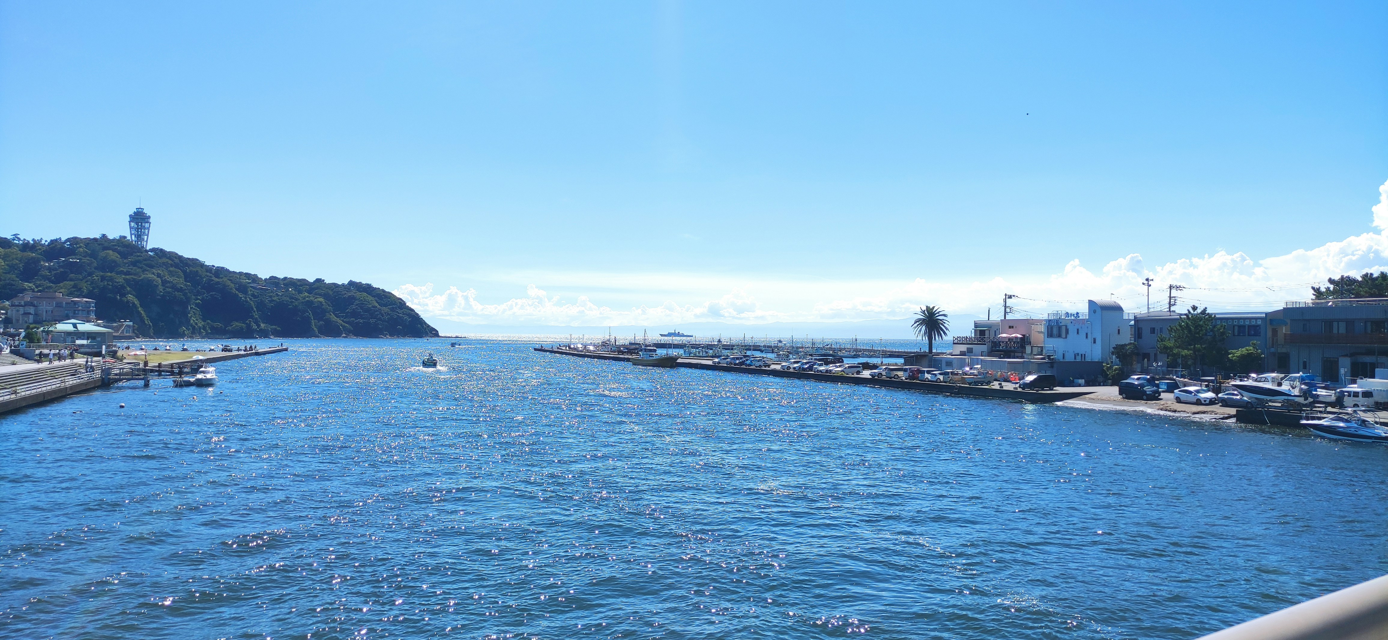 Wide waterway leading to open sea bordered by distant hills and coastal town under clear blue sky.