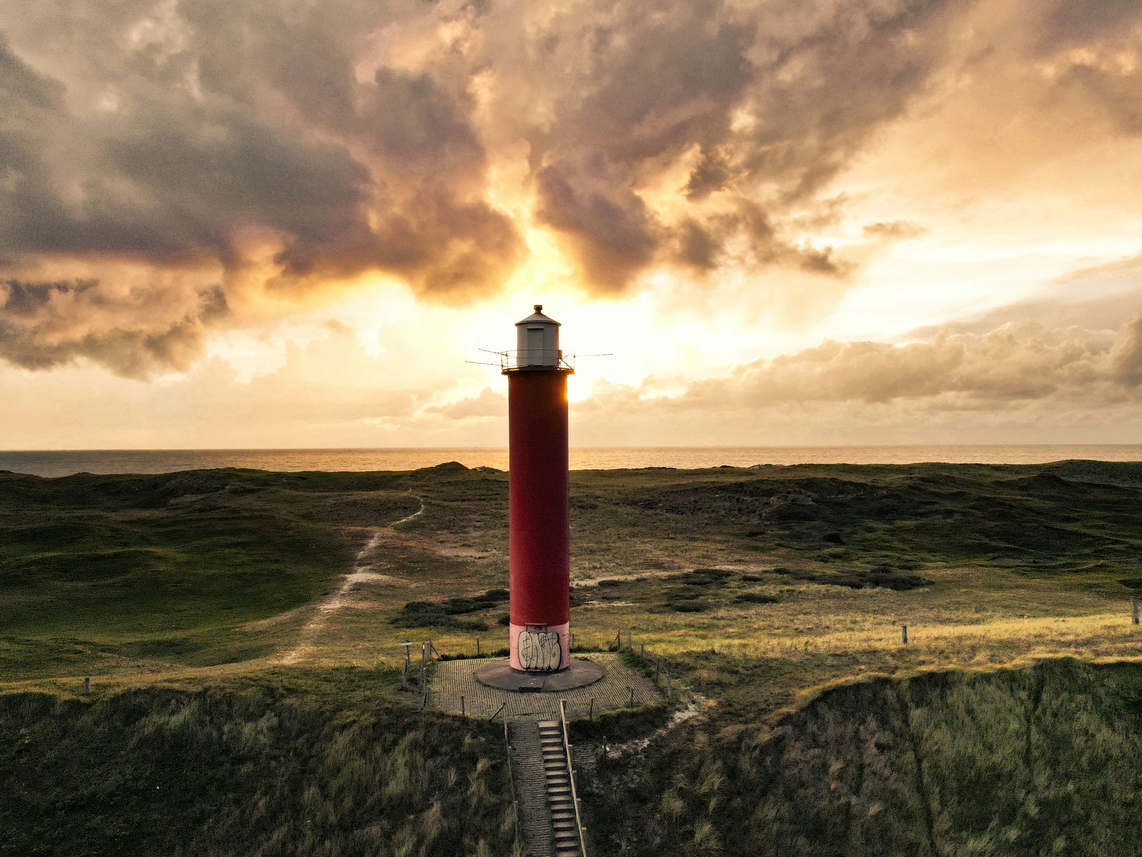 A vibrant red lighthouse stands tall against a dramatic sky, surrounded by rolling dunes and the sea. The scene captures the essence of coastal resilience.