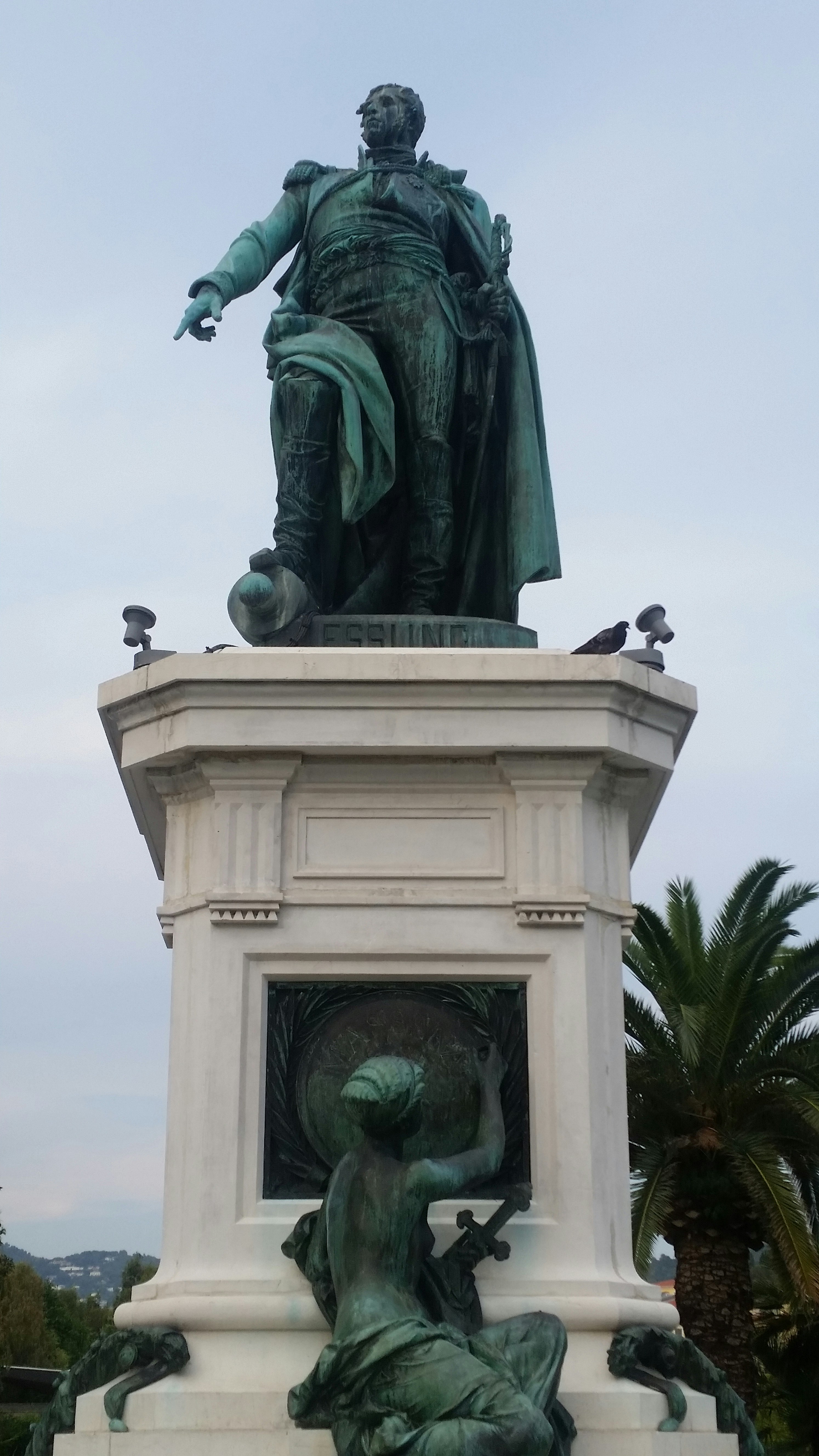Bronze statue of a historical figure standing atop a pedestal, flanked by a seated figure and palm trees in the background.