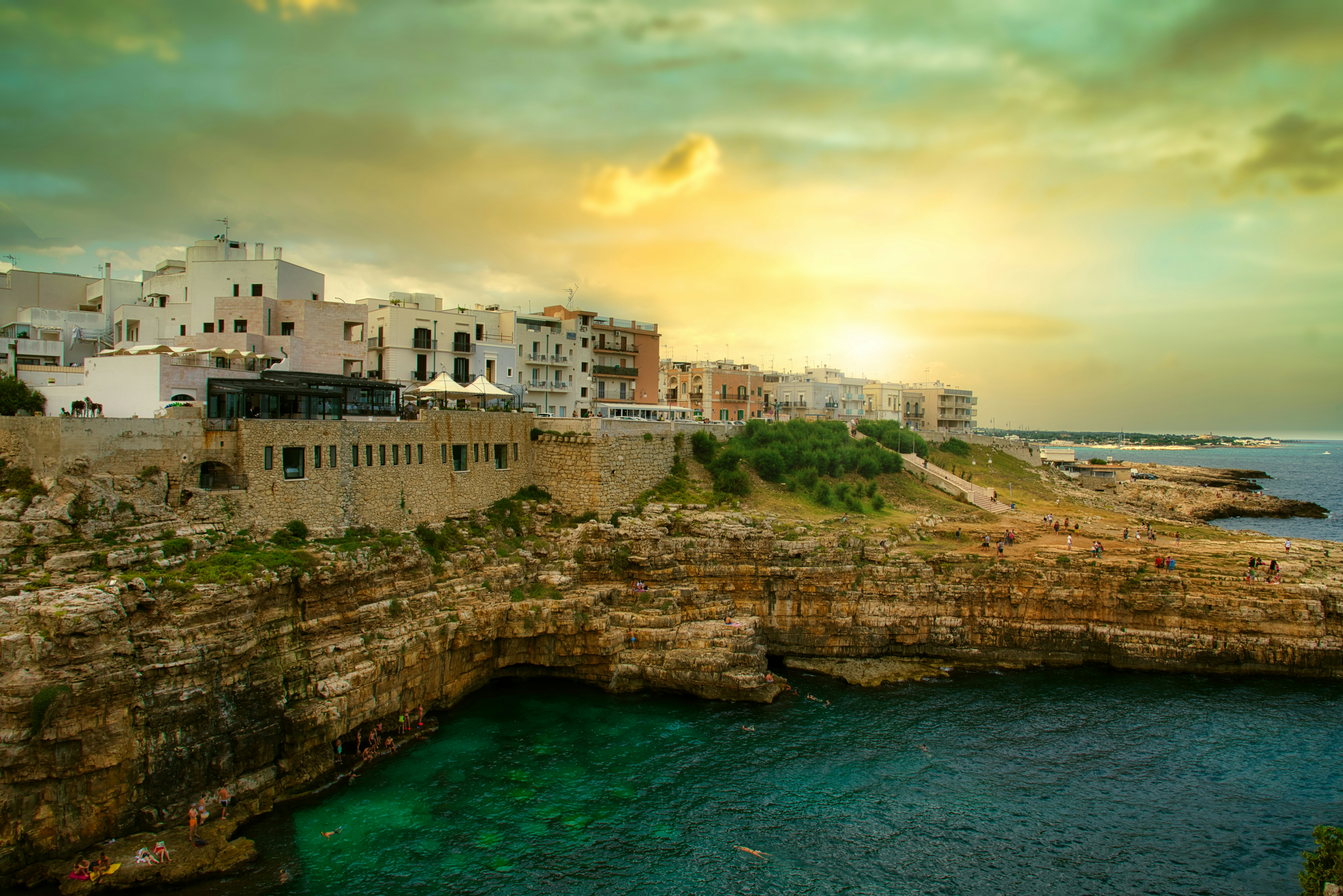 White concrete buildings perched on a cliff overlooking a turquoise sea under a vibrant sky.