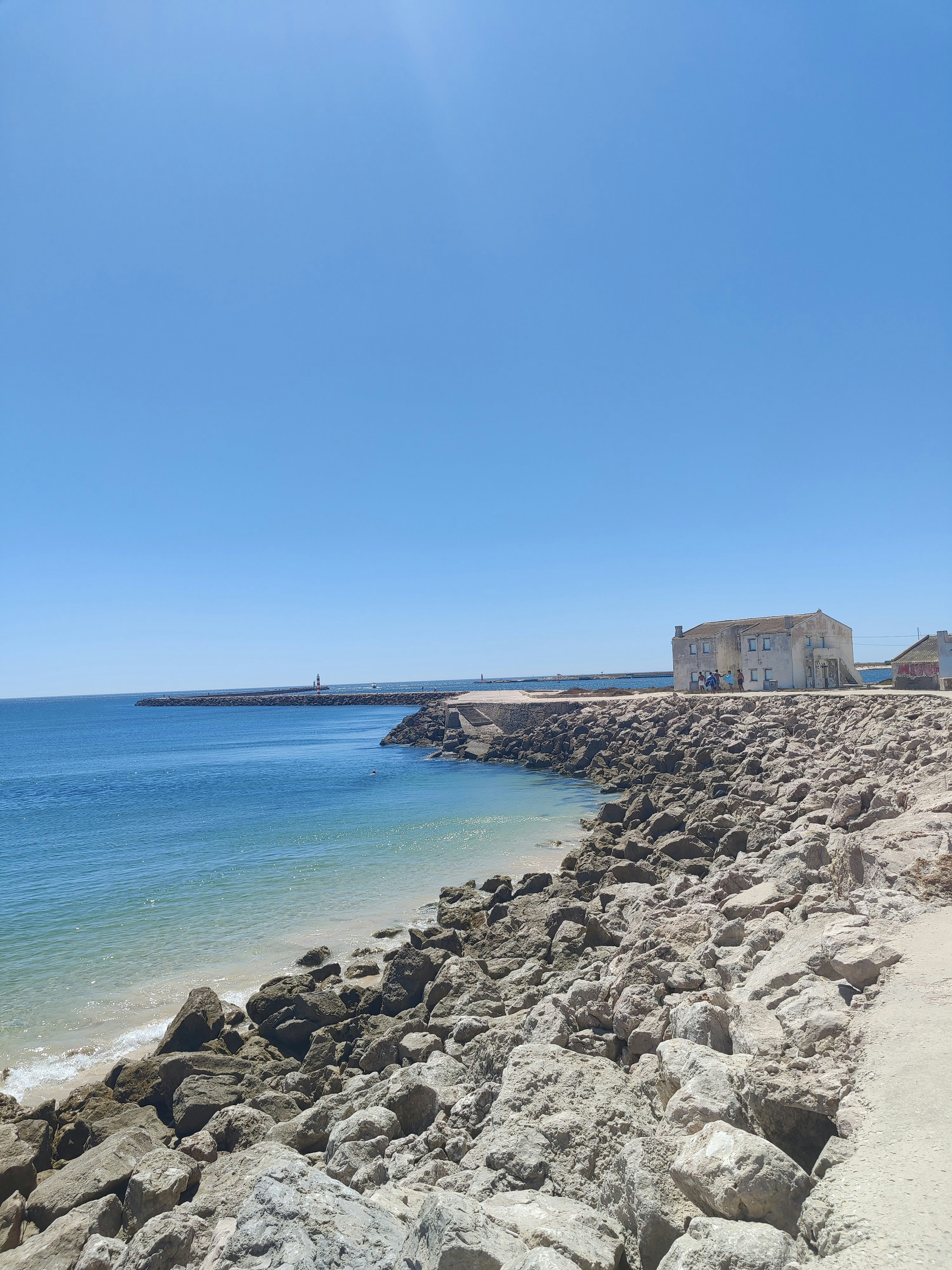Clear seascape with a rocky breakwater leading to a weathered building by calm turquoise waters under a blue sky. A rides along the shore highlights the harbor edge.