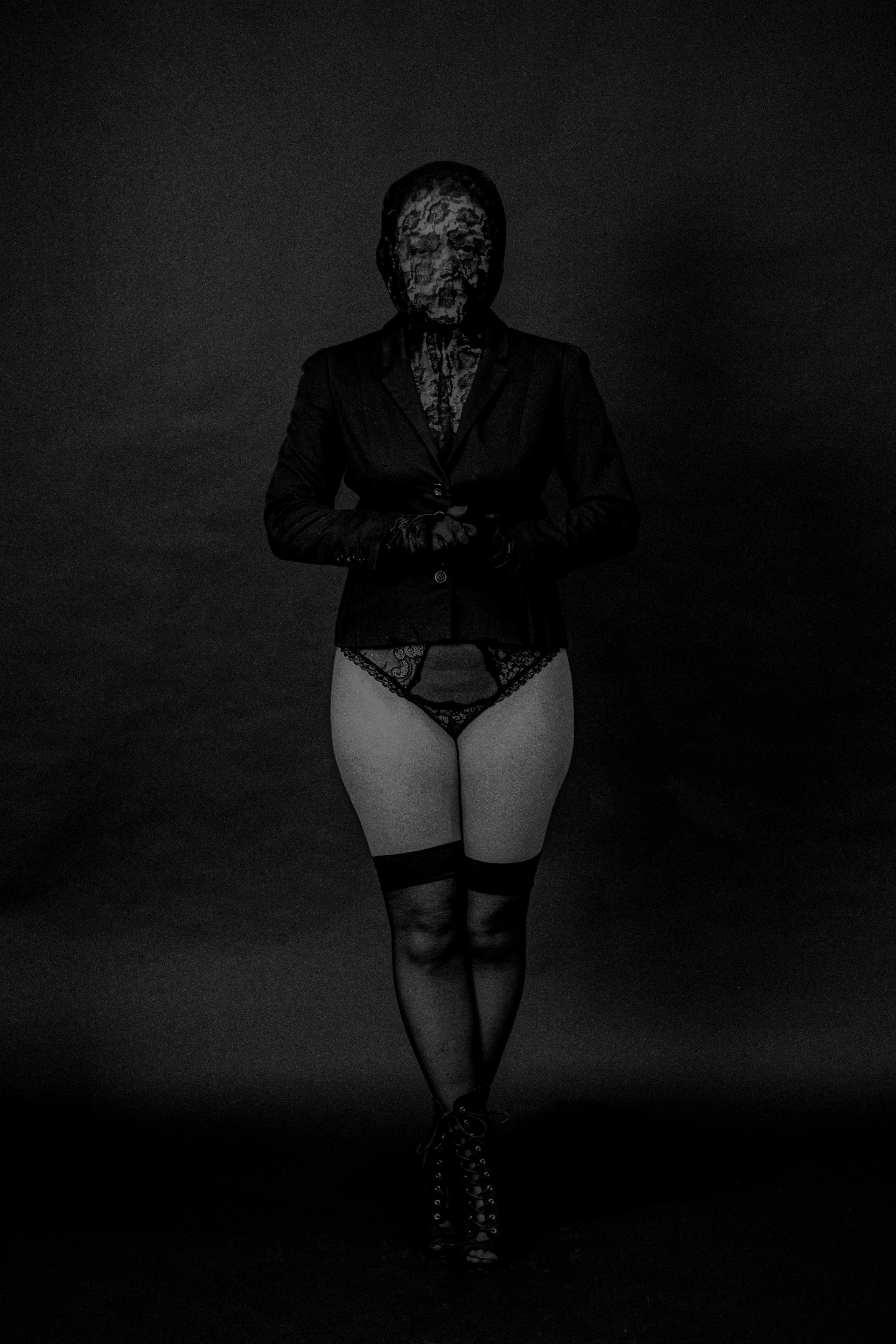 A figure shrouded in a lace mask and formal attire stands against a dark backdrop, merging elegance with mystery.