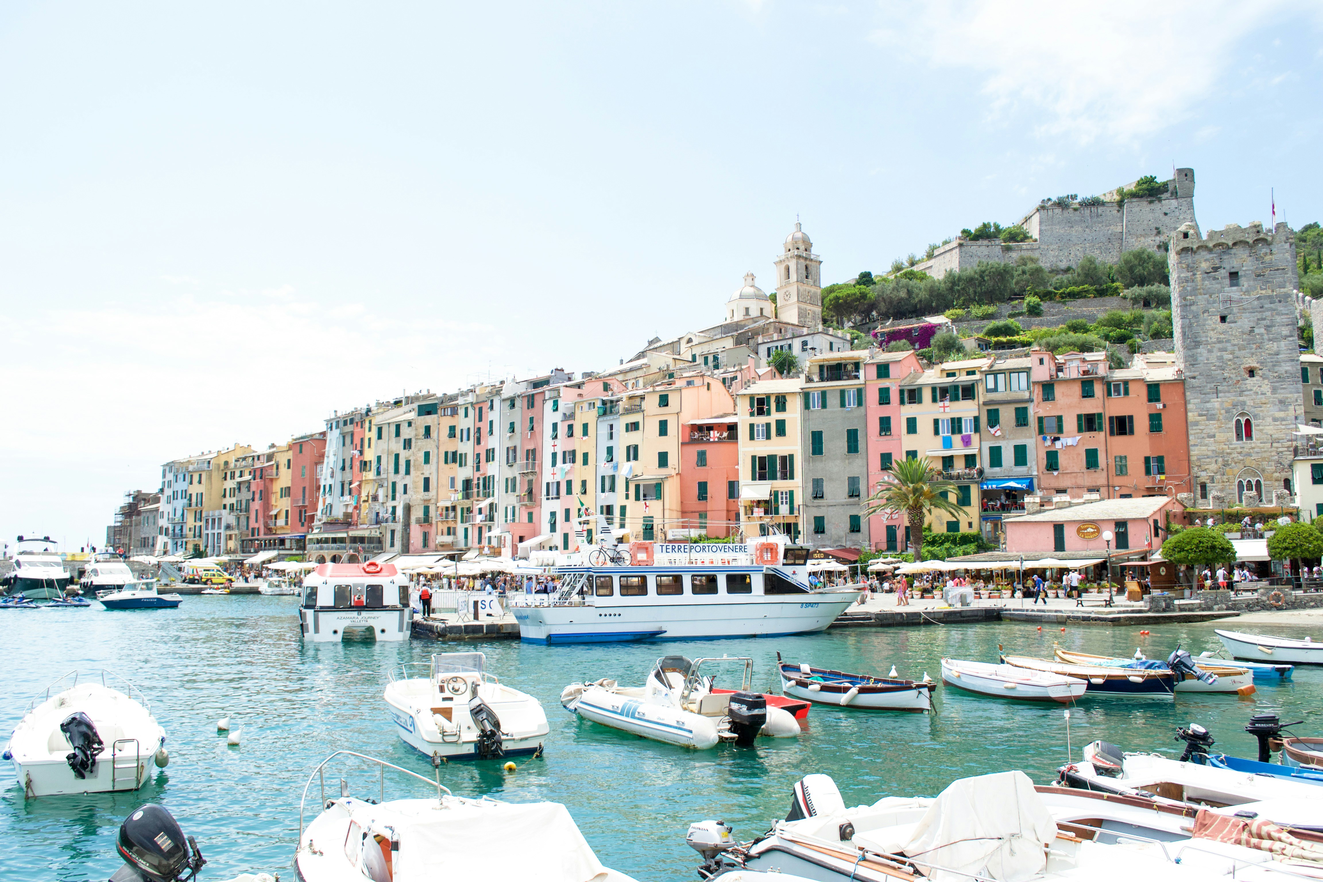 Vibrant harbor scene featuring colorful buildings lining the waterfront, with boats gently bobbing in the clear waters. A historic fortress looms in the background.