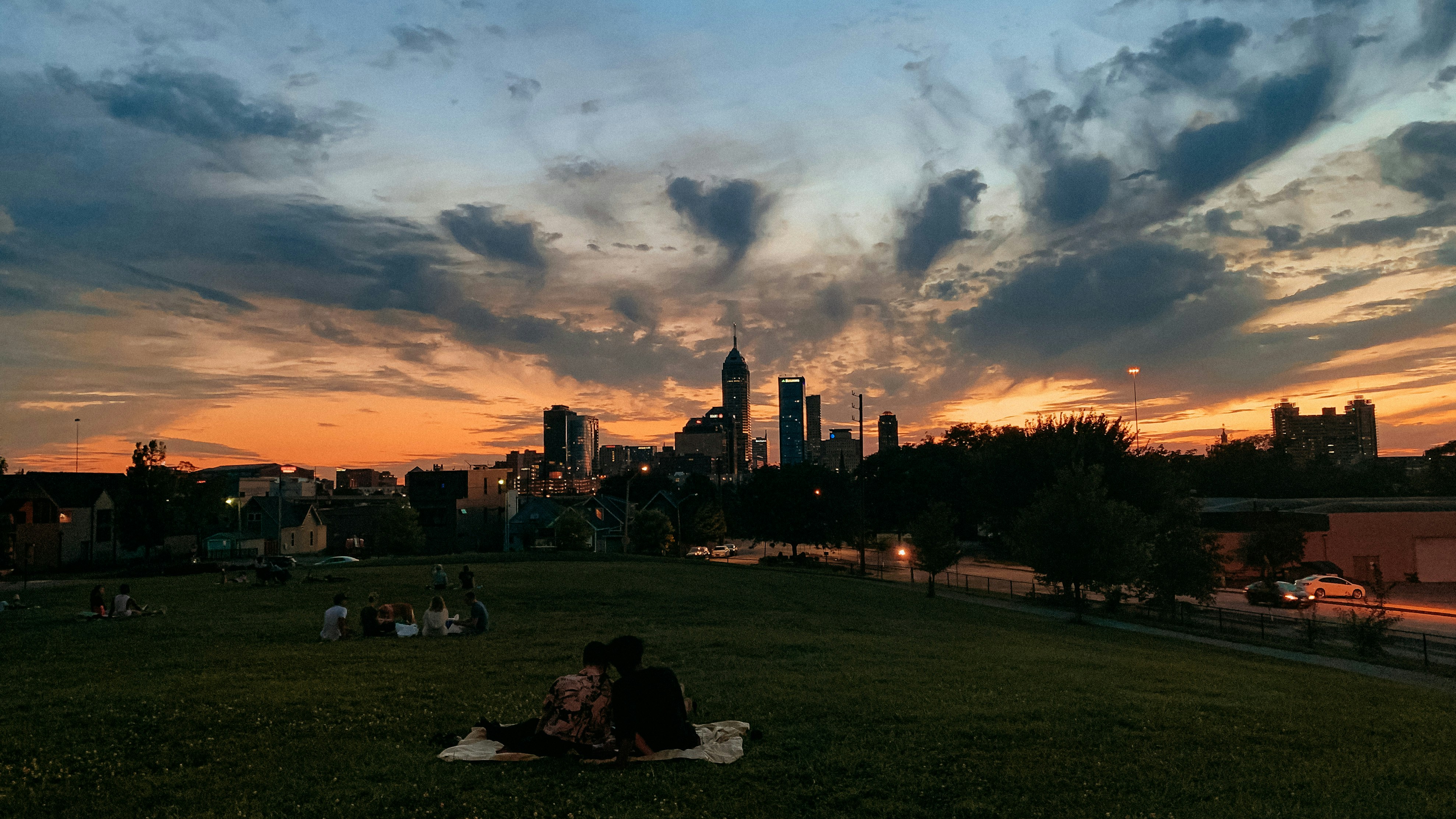 People relaxing on a grassy field as the city skyline is silhouetted against a vibrant sunset.