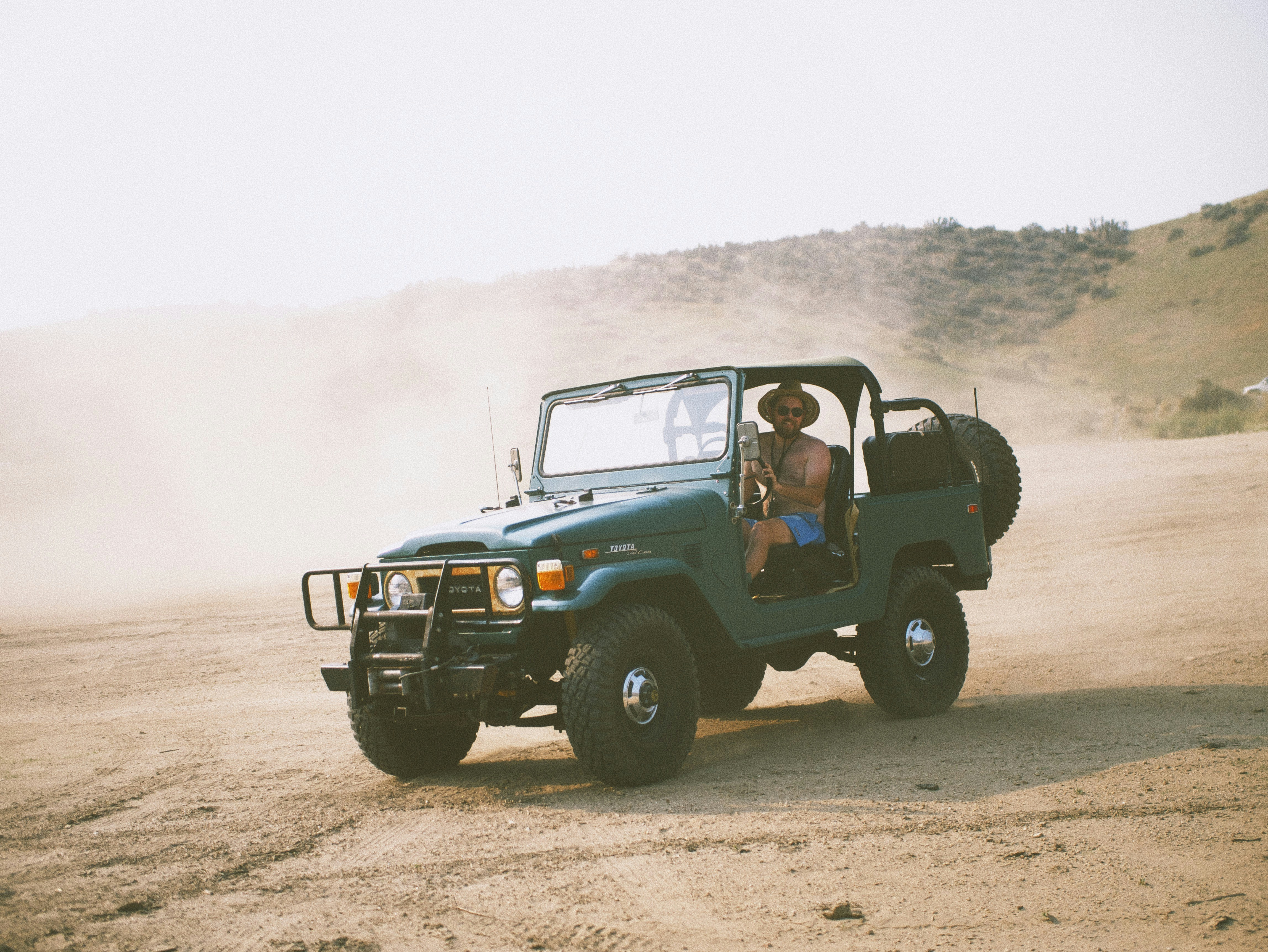Vintage green Jeep Wrangler navigating a dusty landscape with hills in the background.
