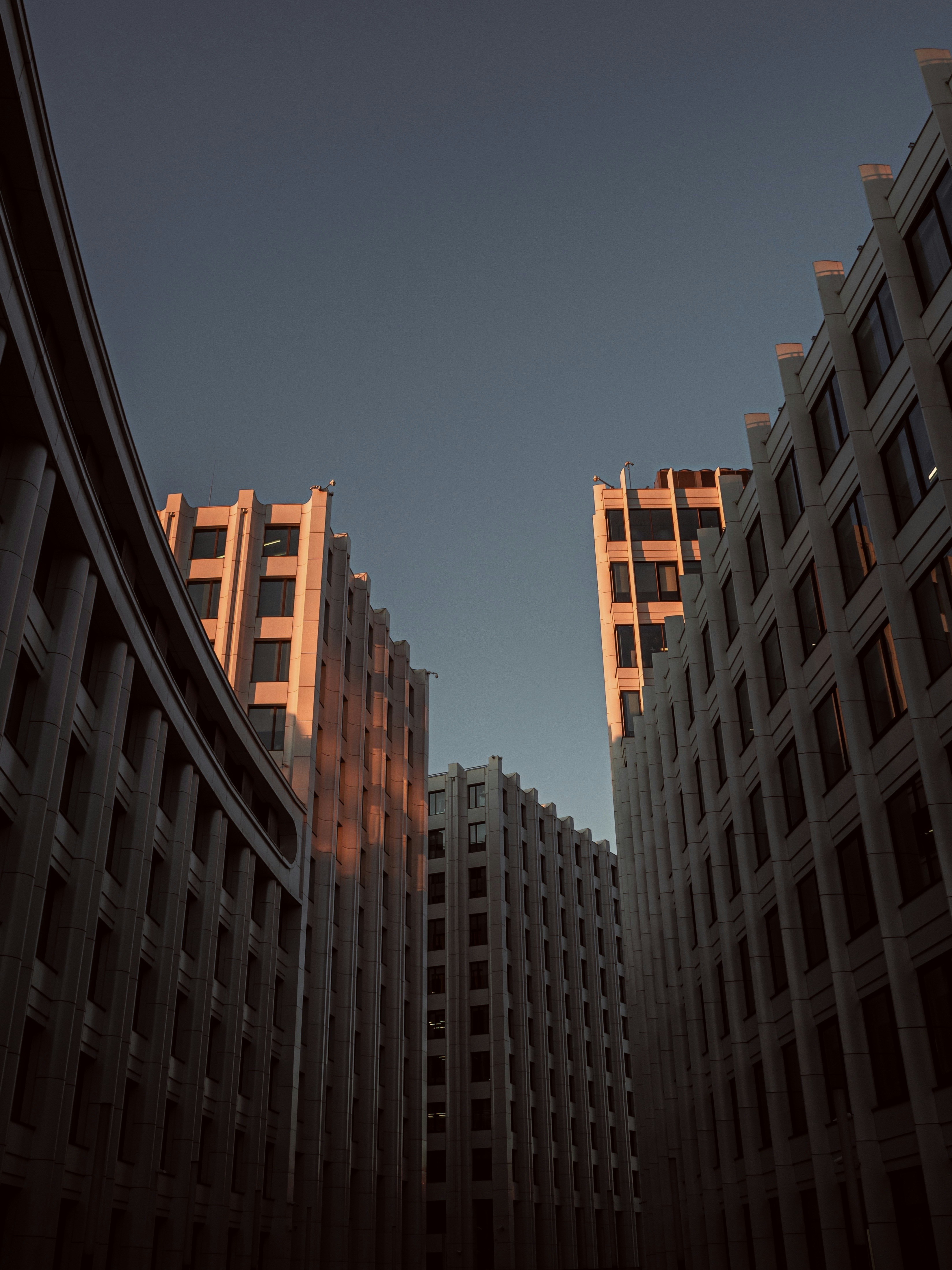 Tall modern buildings with angular designs illuminated by the warm glow of sunset, creating a striking contrast against the twilight sky.