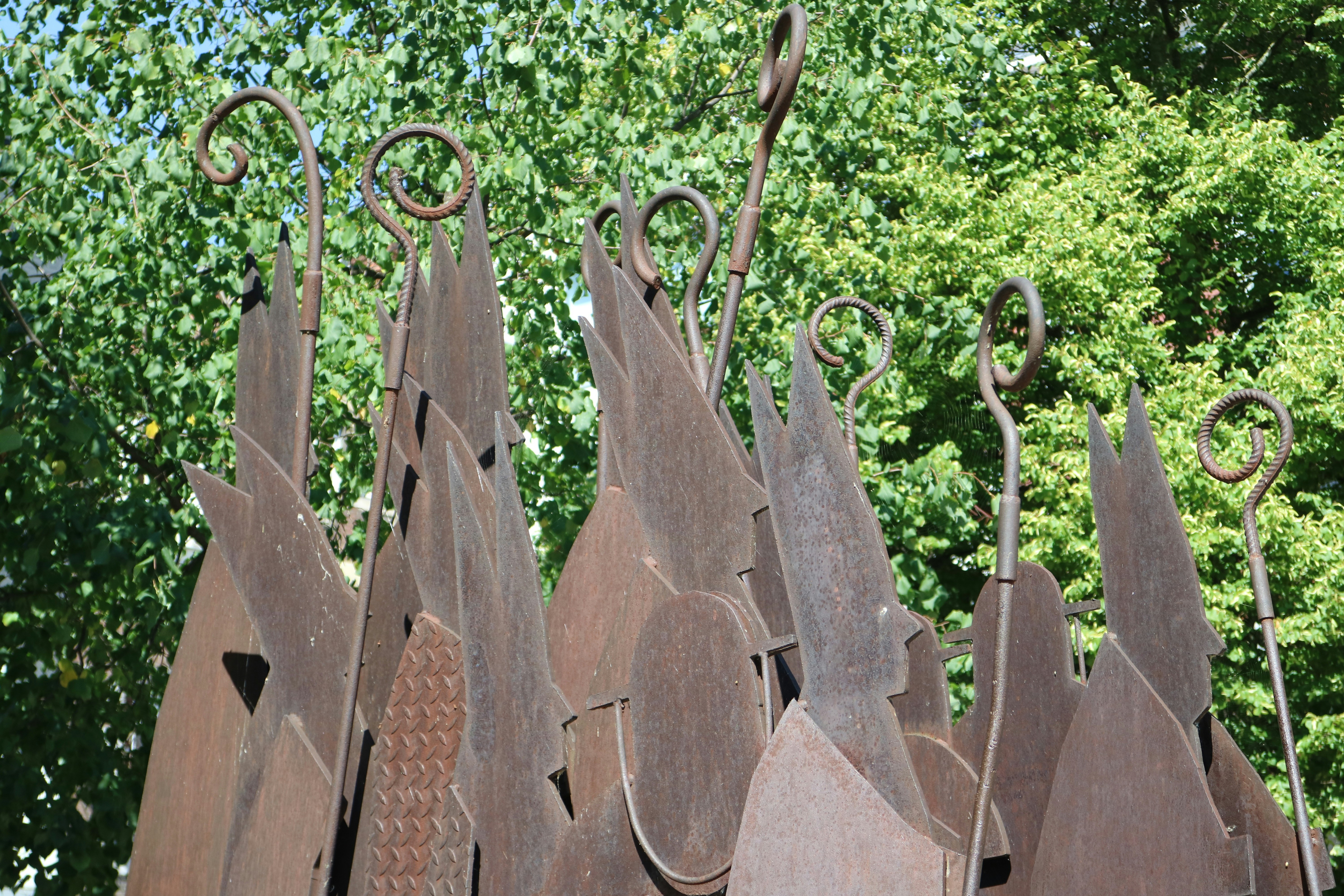 Rust-colored metal sculptures with pointed tops set against a backdrop of lush green trees.