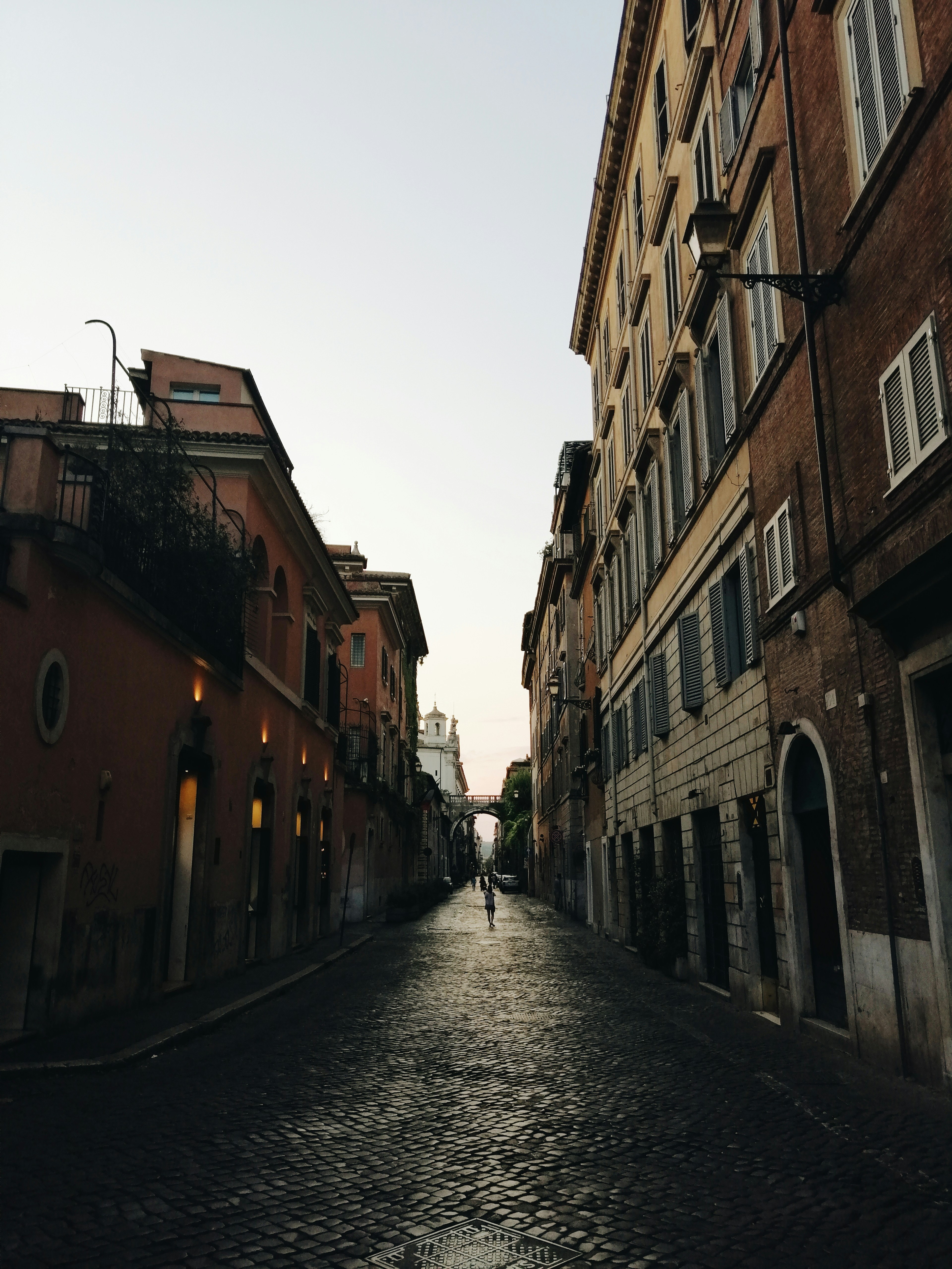 Narrow cobblestone street in Rome at dusk, lined with historic buildings and soft glowing lights. A solitary figure walks towards an archway in the distance.