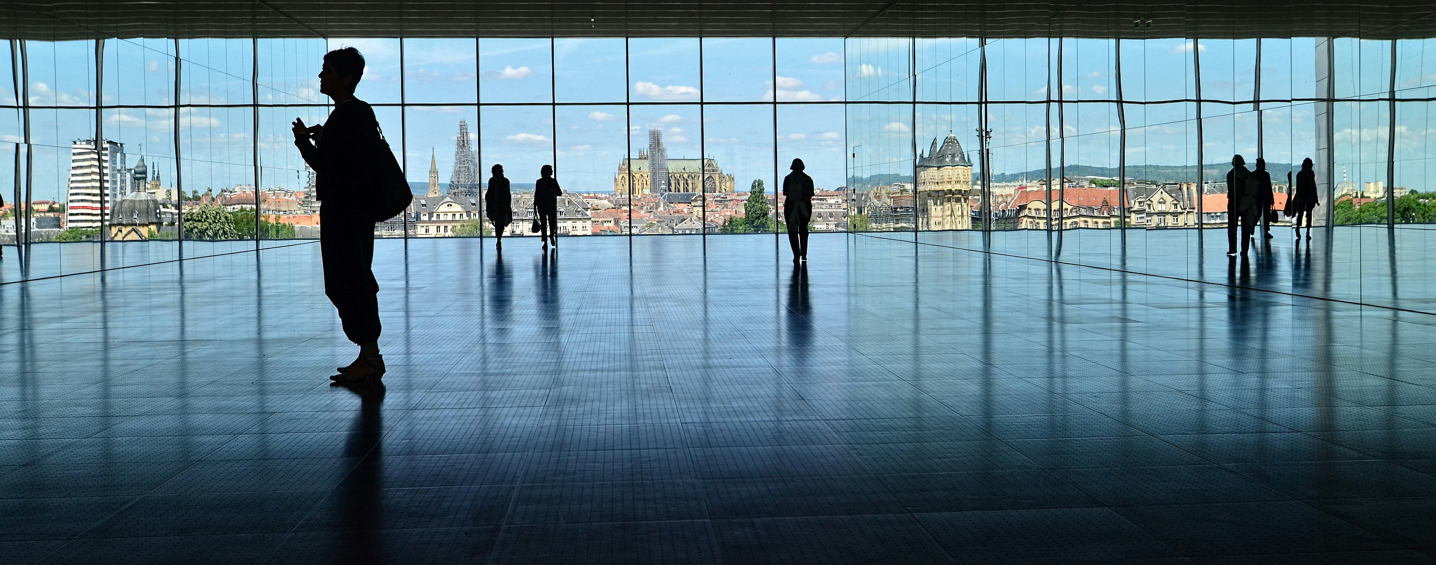 Silhouetted figures walk along a glossy floor with a cityscape reflected in large windows.
