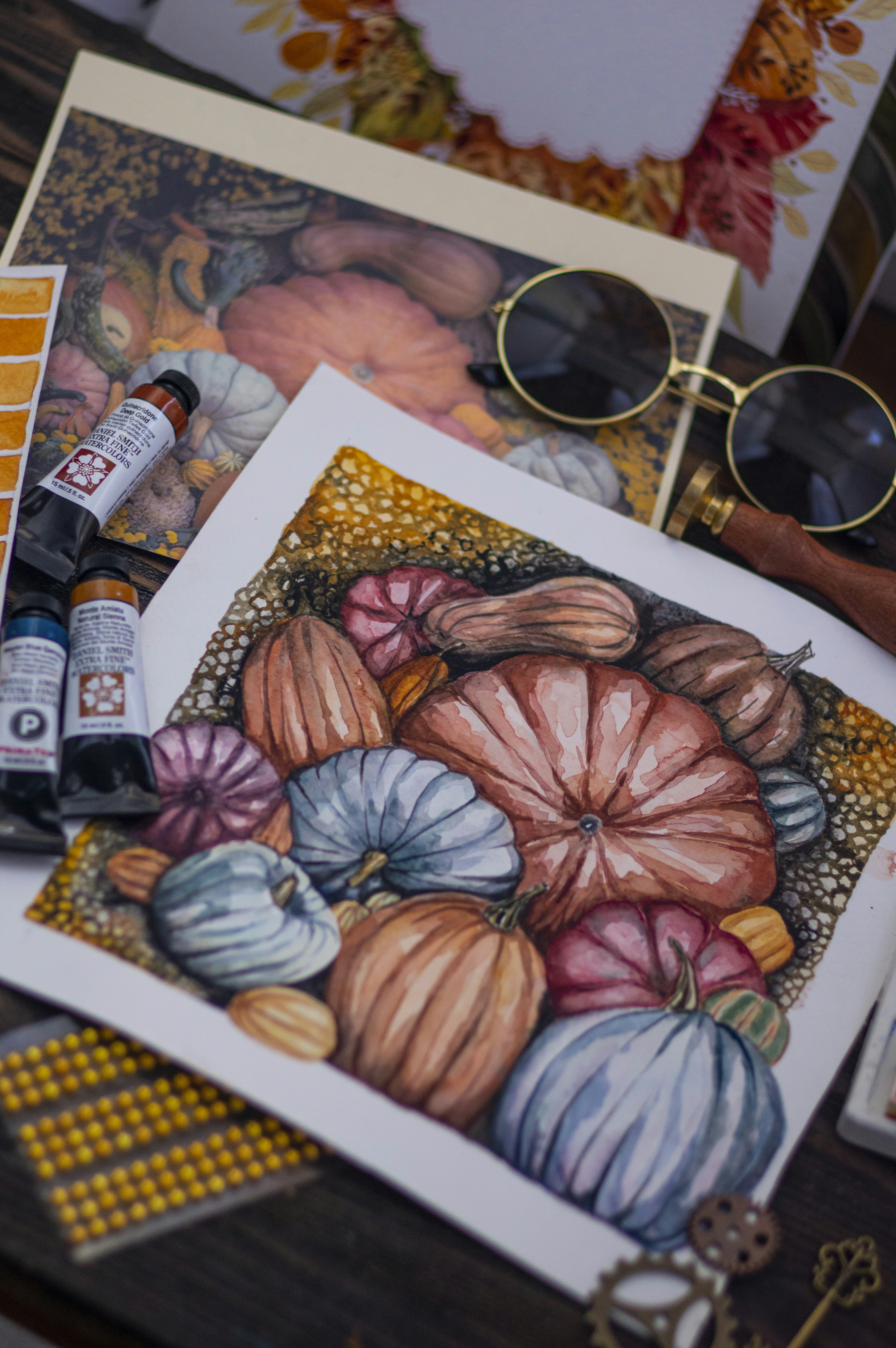 Colorful pumpkin painting laid atop printed autumn patterns, surrounded by paint tubes, beads, and vintage sunglasses. A still-life that emphasizes warm hues, texture, and a cozy craft workspace.
