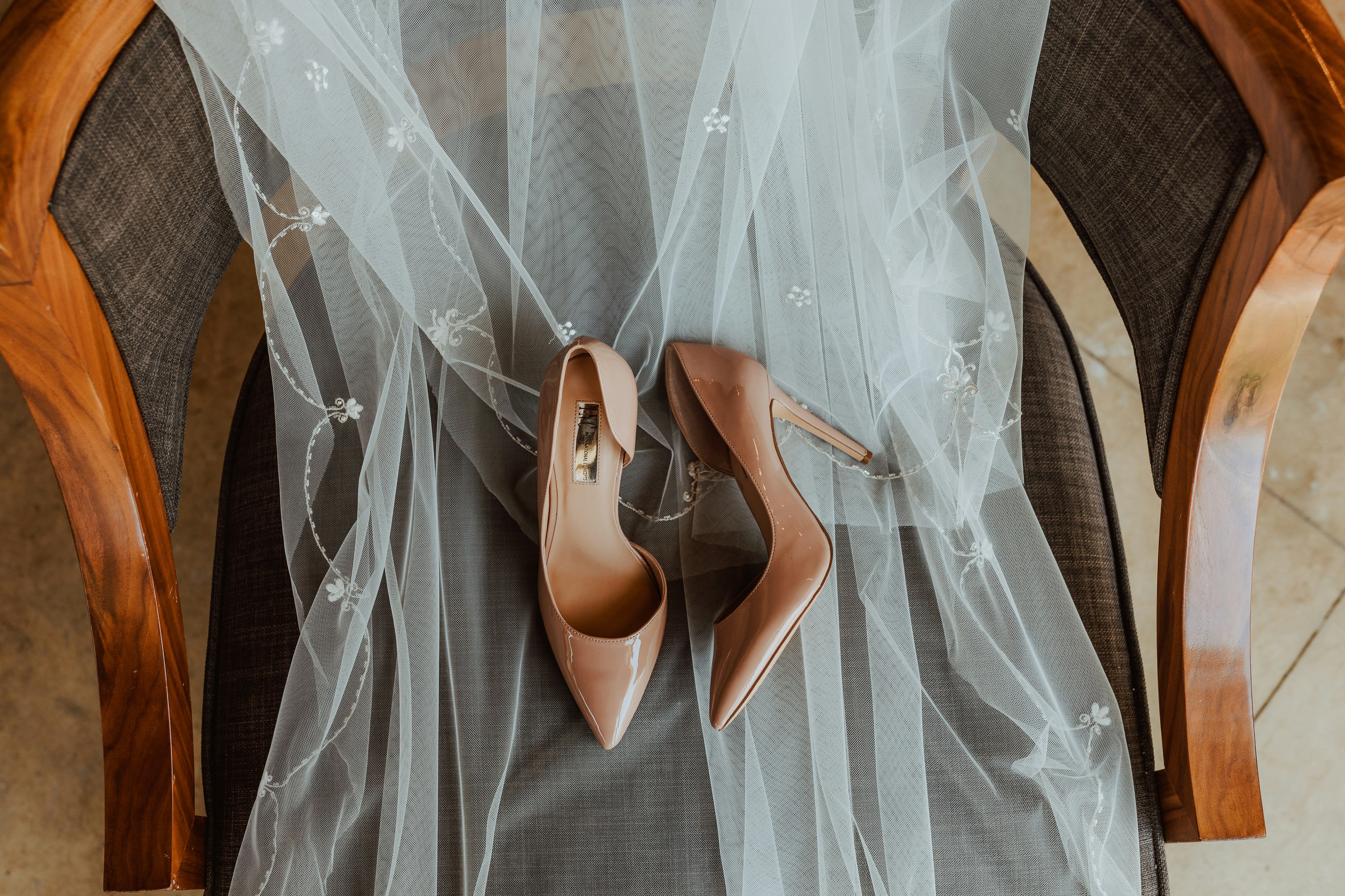 A pair of nude high heels resting on a textured chair, draped with delicate bridal veil. The soft fabric and polished shoes create a serene bridal atmosphere.