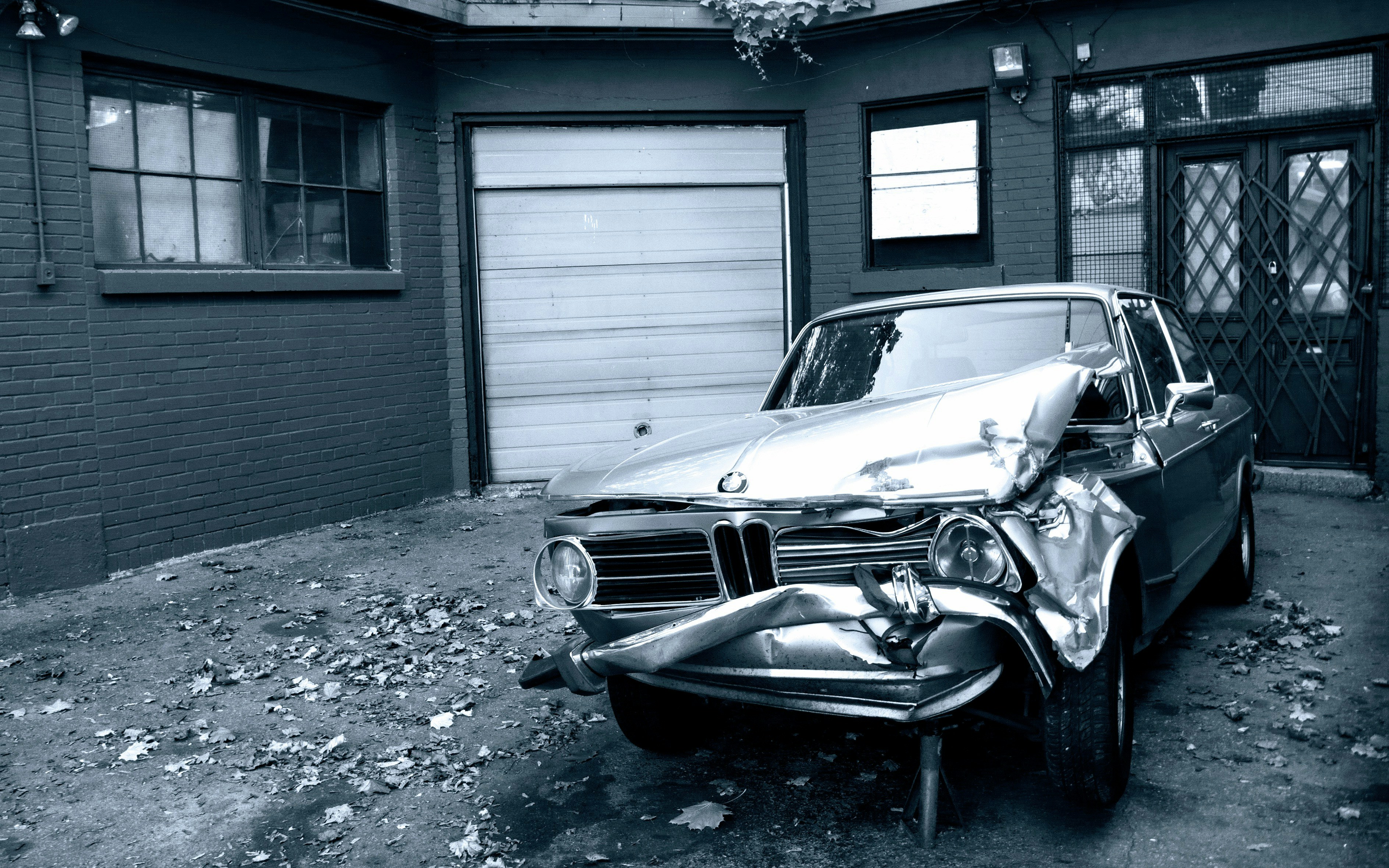 Monochrome photograph of a damaged vintage sedan in a gravel courtyard, its front end crumpled as a garage door and brick walls frame the scene.