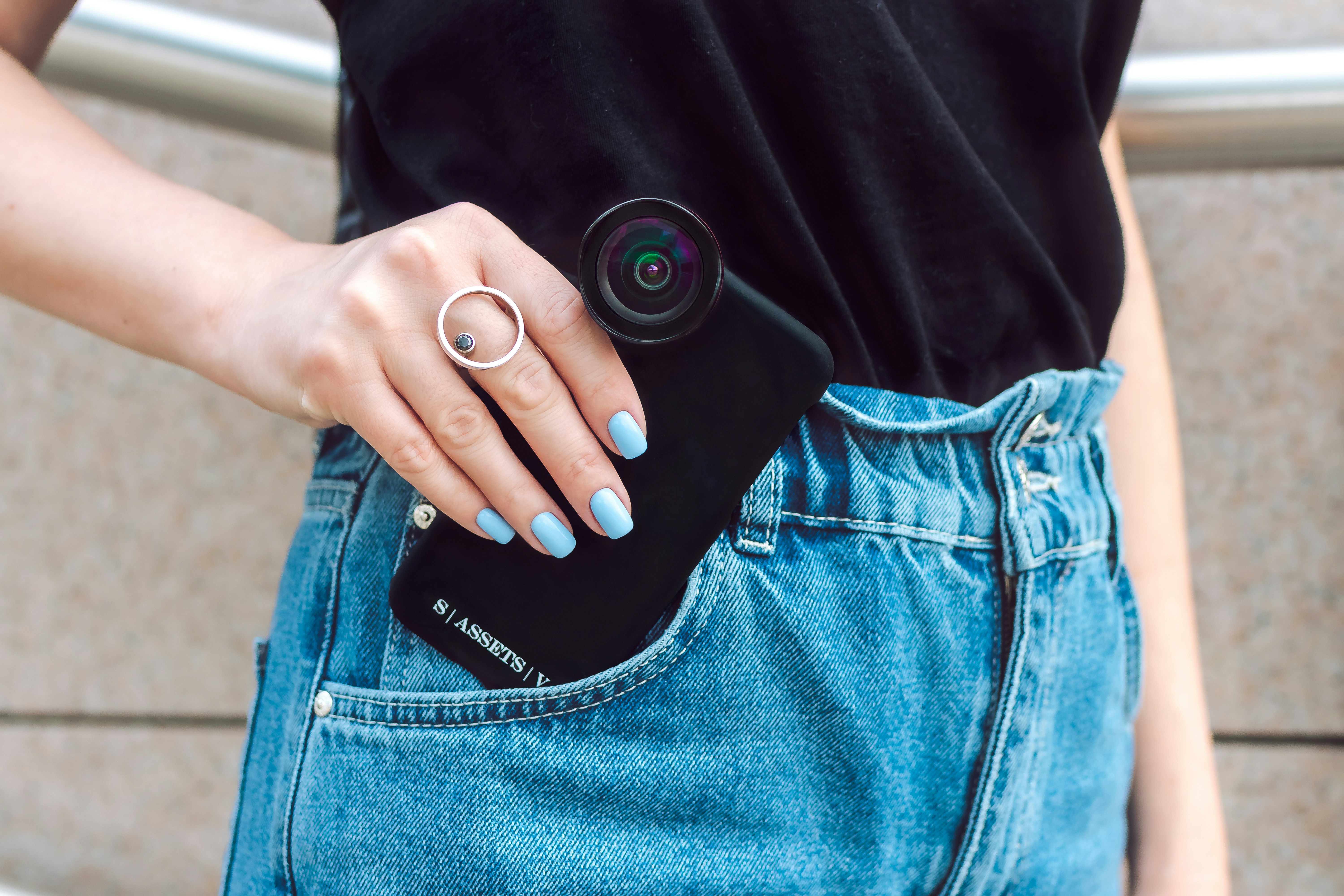 A hand holding a smartphone equipped with a lens attachment, tucked into high-waisted denim shorts against a textured background.