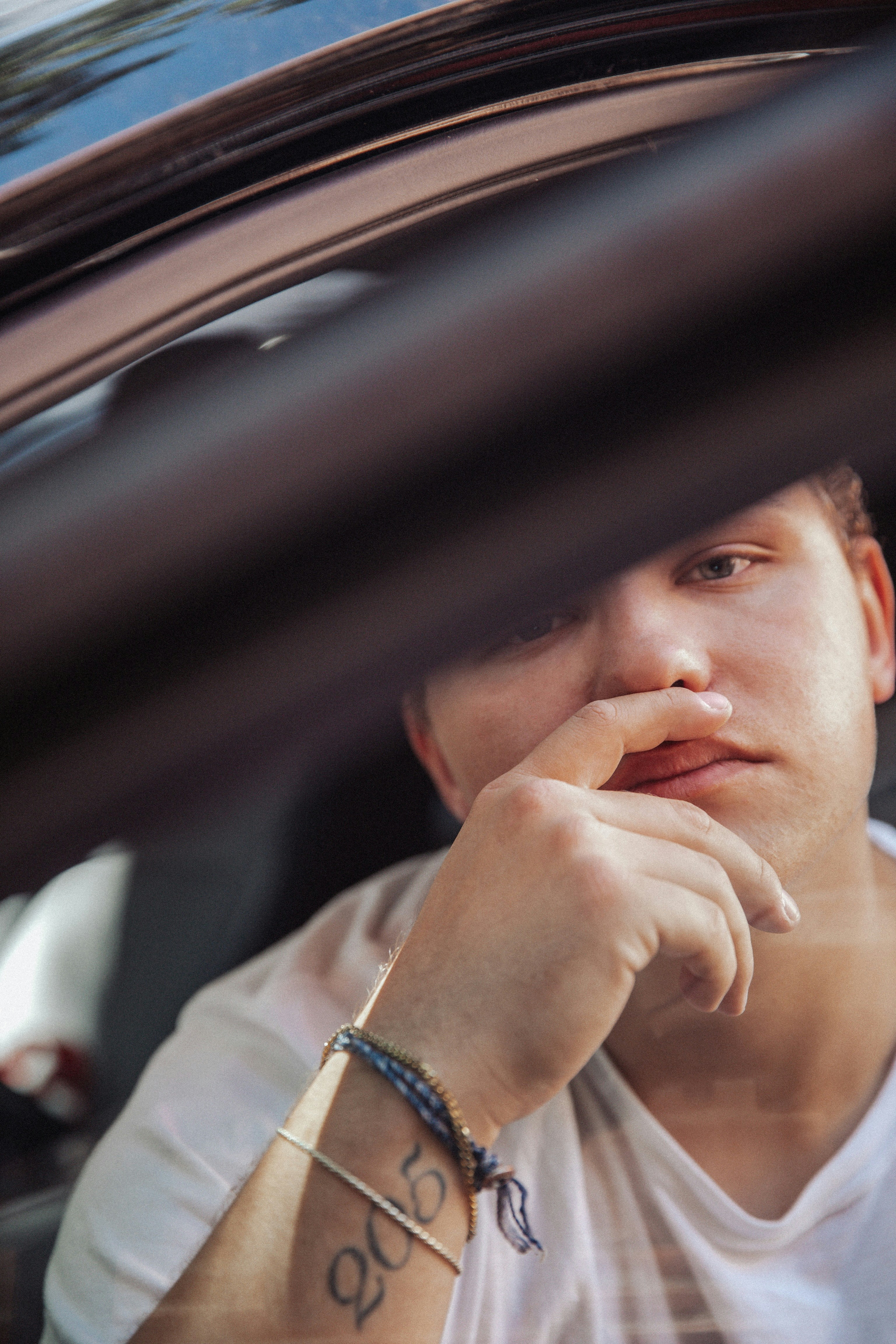 Young man gazing thoughtfully from a car window, hand resting on his chin, with sunlight filtering through the glass.