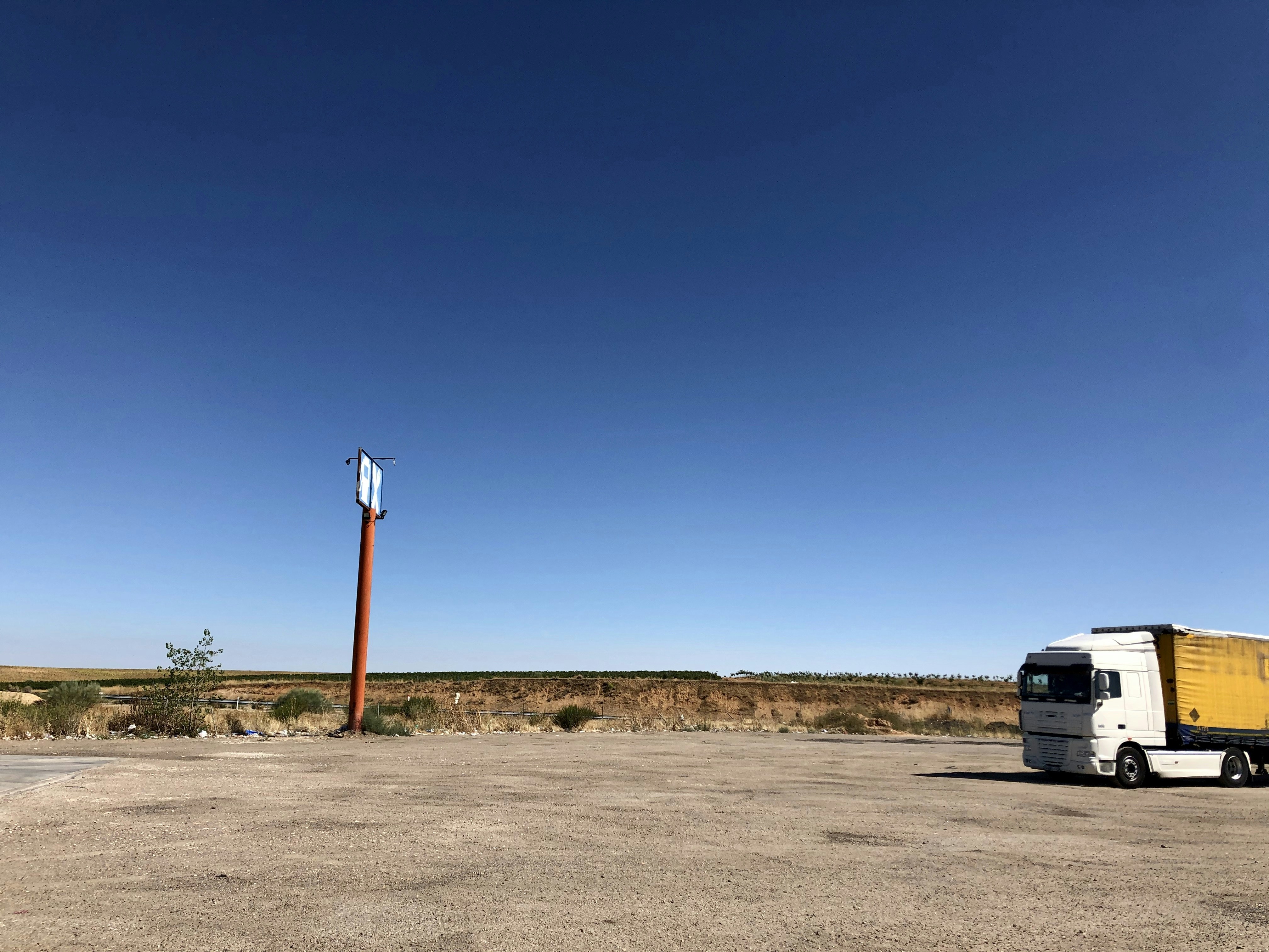 A parked truck rests on an expansive, empty lot under a clear blue sky, with a lone signpost in the background. The scene captures the essence of solitude in a wide-open landscape.