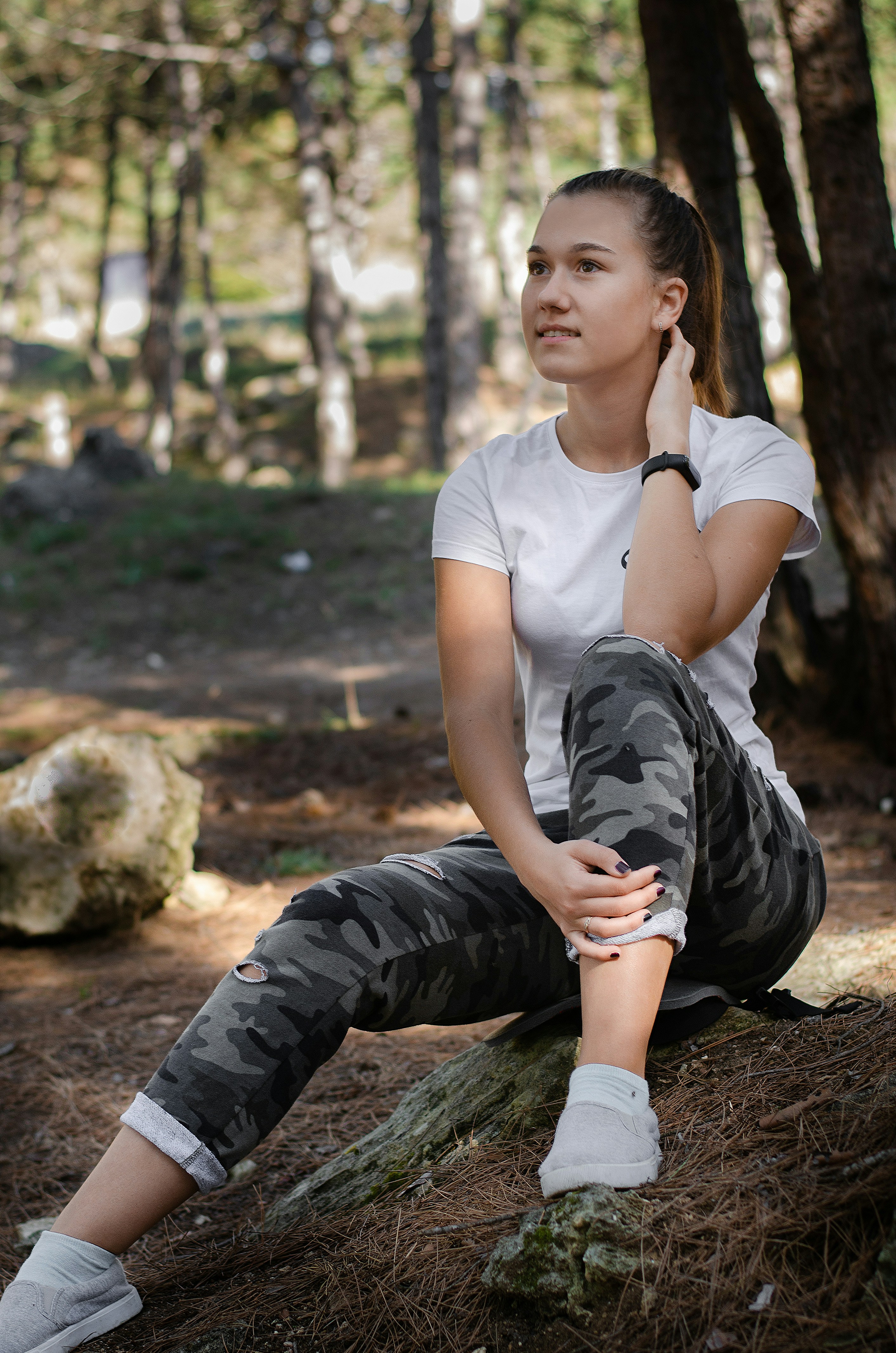 Young woman seated on a rock, gently touching her hair, surrounded by trees and natural elements. The sunlight filters through the foliage, creating a serene atmosphere.