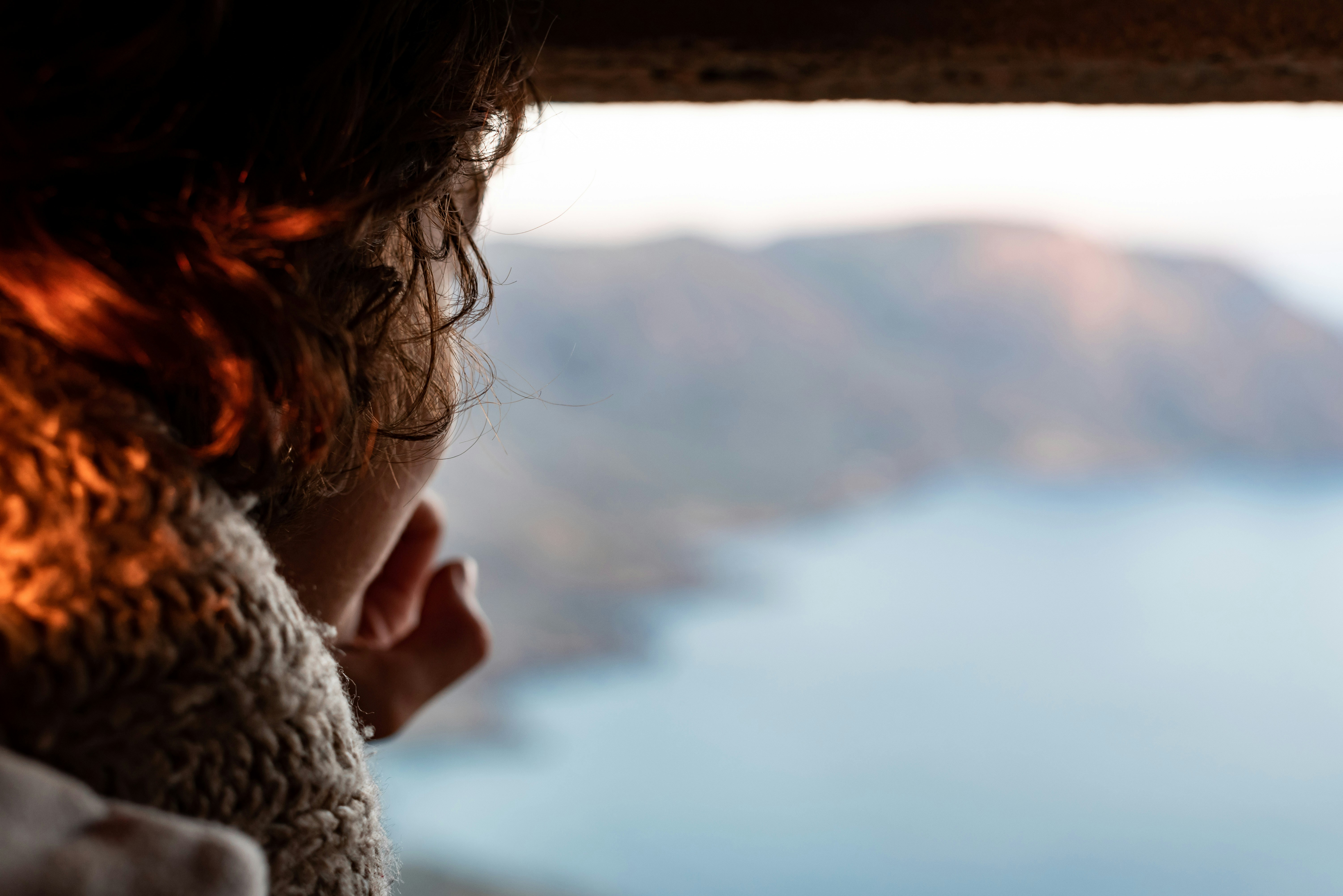 Person gazing out from a window towards a blurred coastal landscape at sunset.